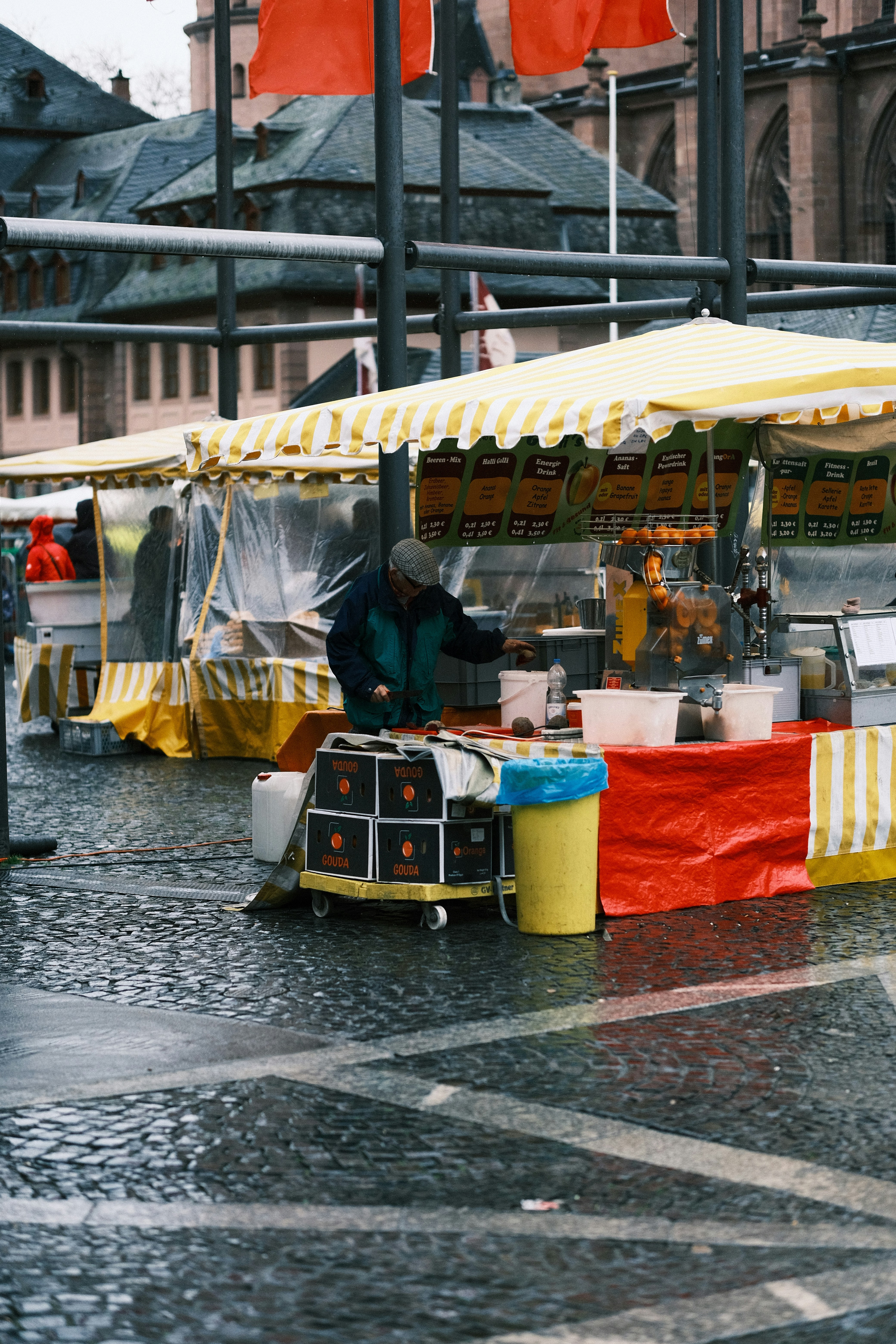 A vendor prepares food at a vibrant market stall, surrounded by colorful awnings and a rainy atmosphere. The scene captures the essence of street food culture.