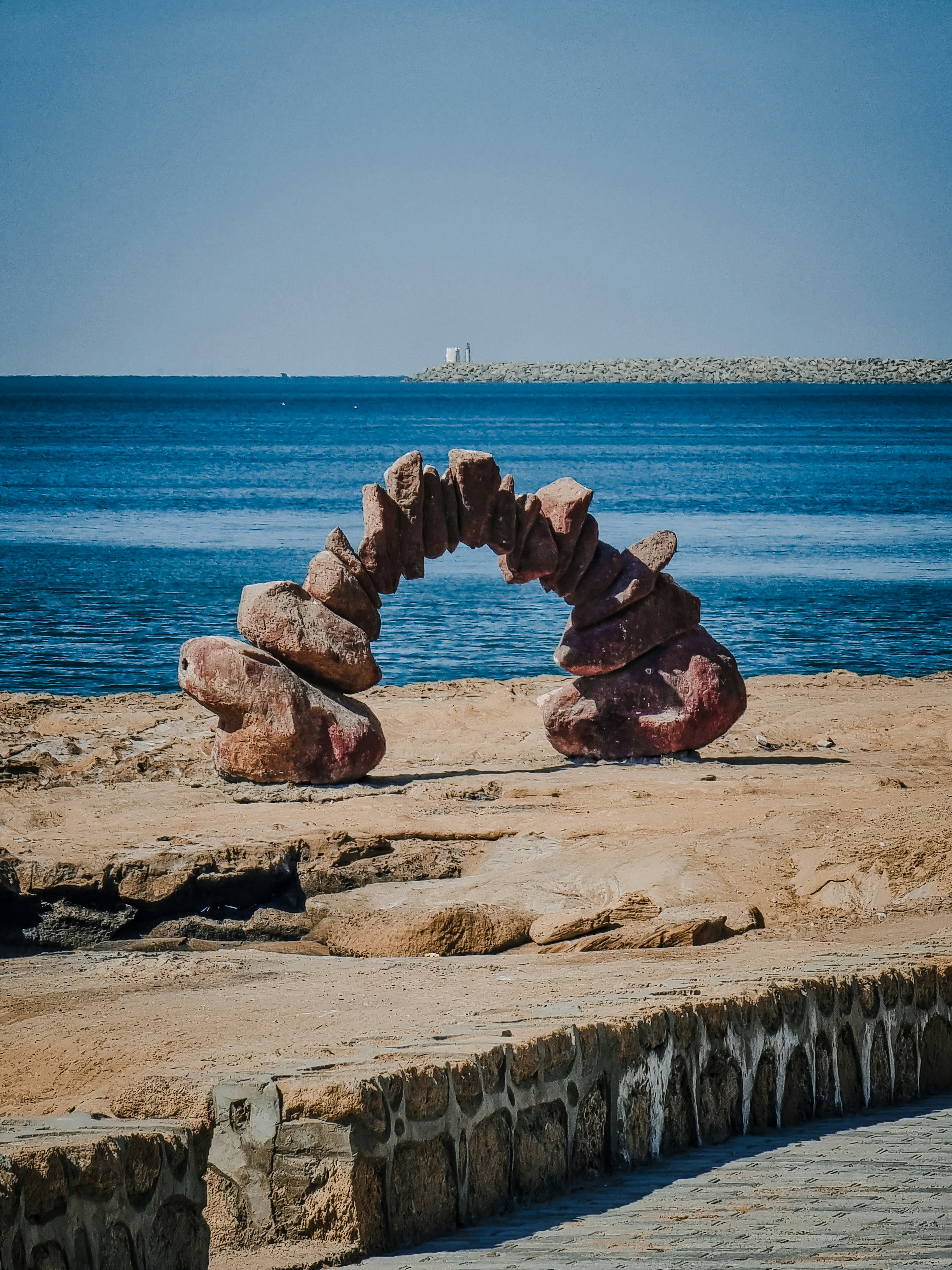 Stacked-stone arch on a sunlit sandy jetty overlooking the blue sea and sky.