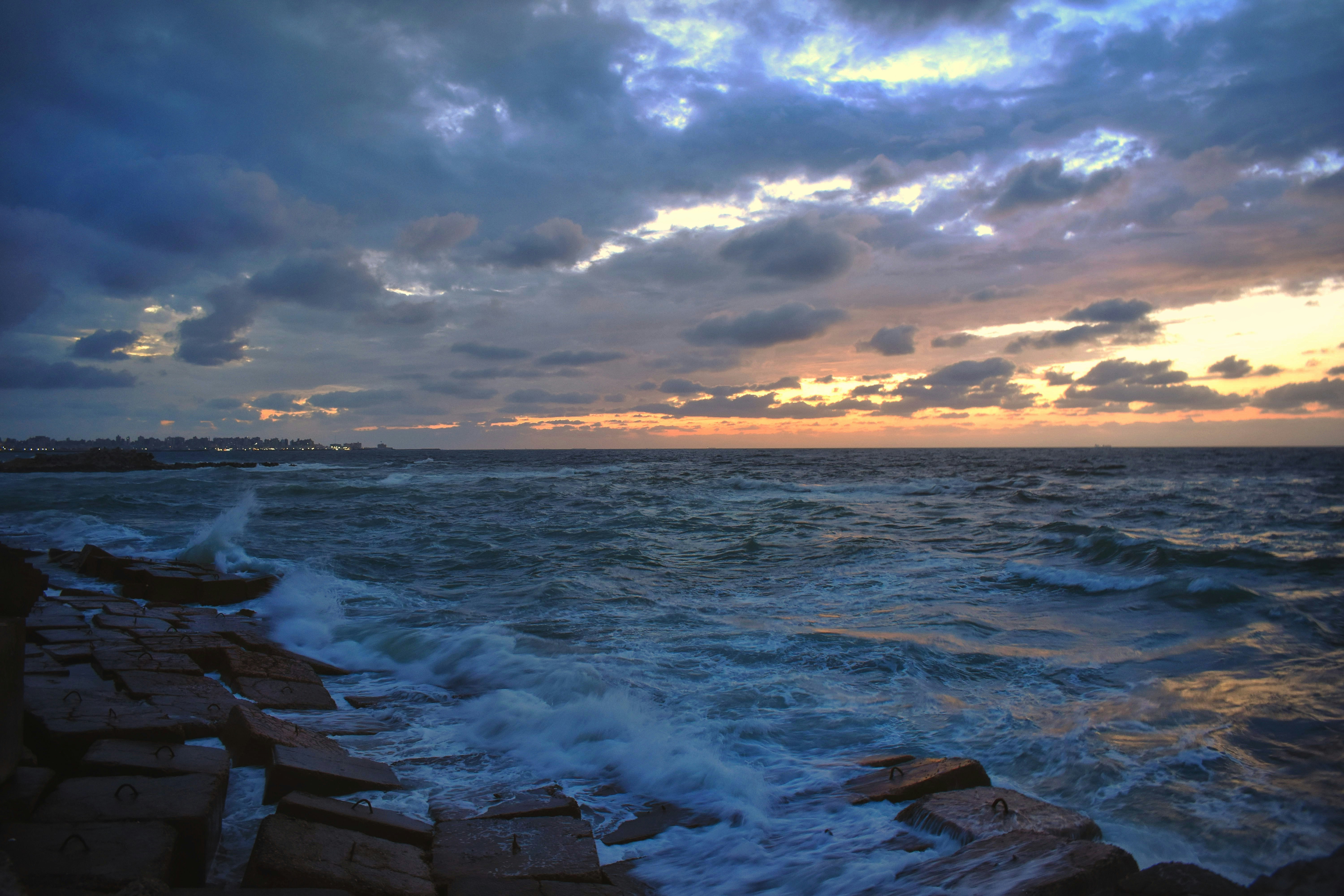 Sunset over the ocean with waves crashing against rugged rocks under a dramatic sky.