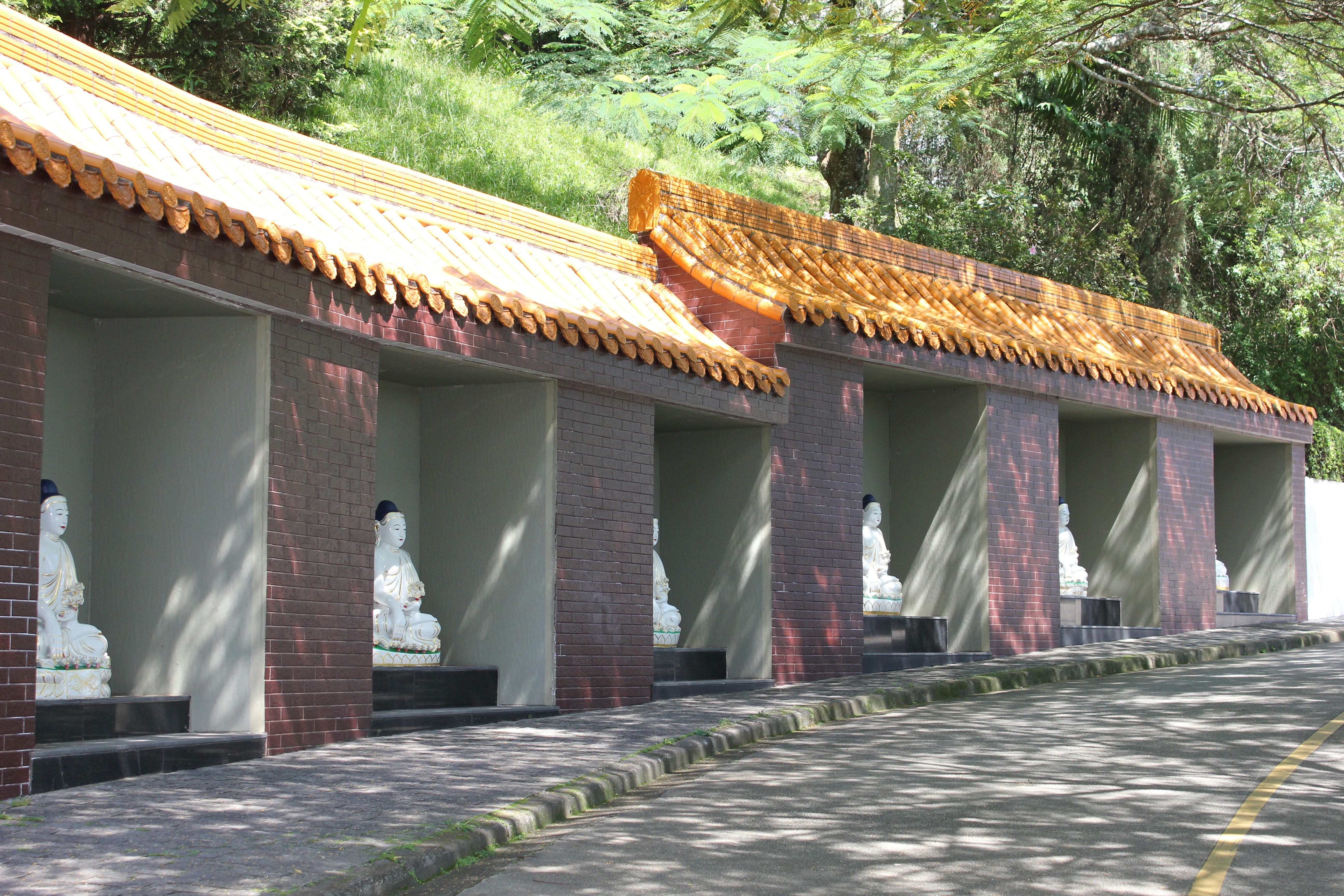 Buddha statues arranged under traditional roofs, lining a winding road through lush greenery.