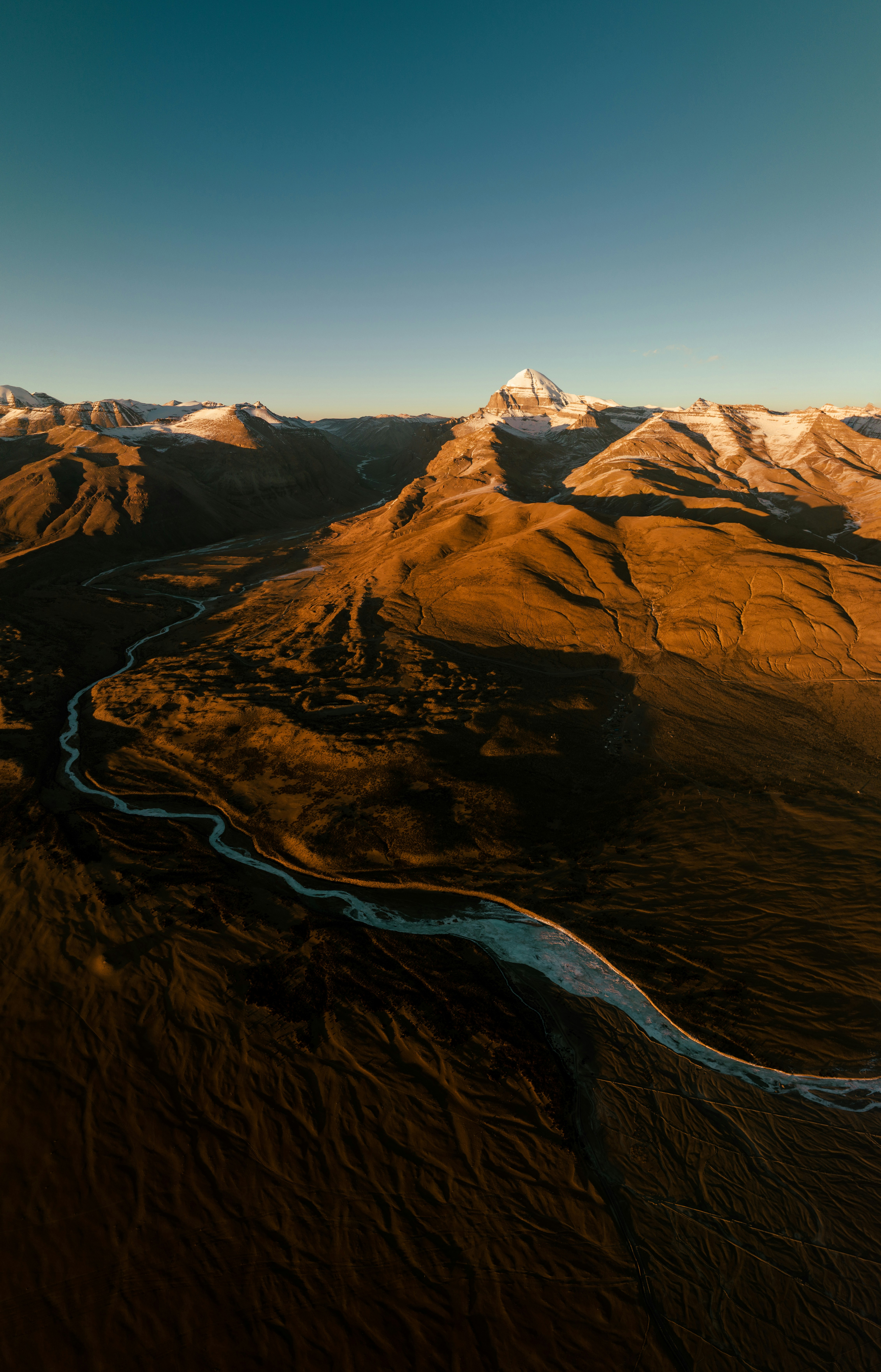 Drone photograph of a snow-capped peak bathed in golden light, with a winding river cutting through rugged, ochre-toned valleys.