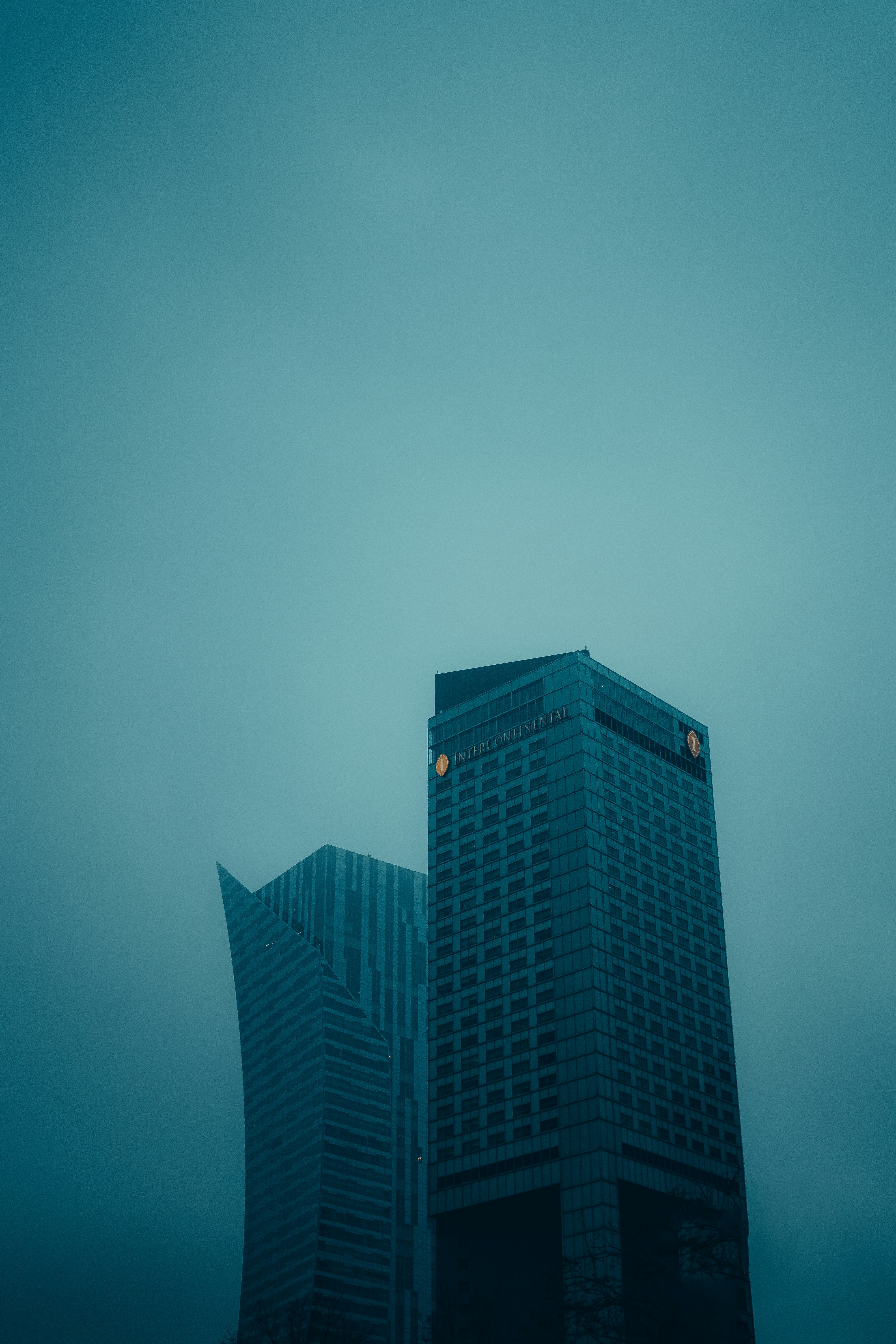Two skyscrapers emerge through a foggy atmosphere, showcasing modern architecture against a muted backdrop. The scene captures a sense of urban mystery.