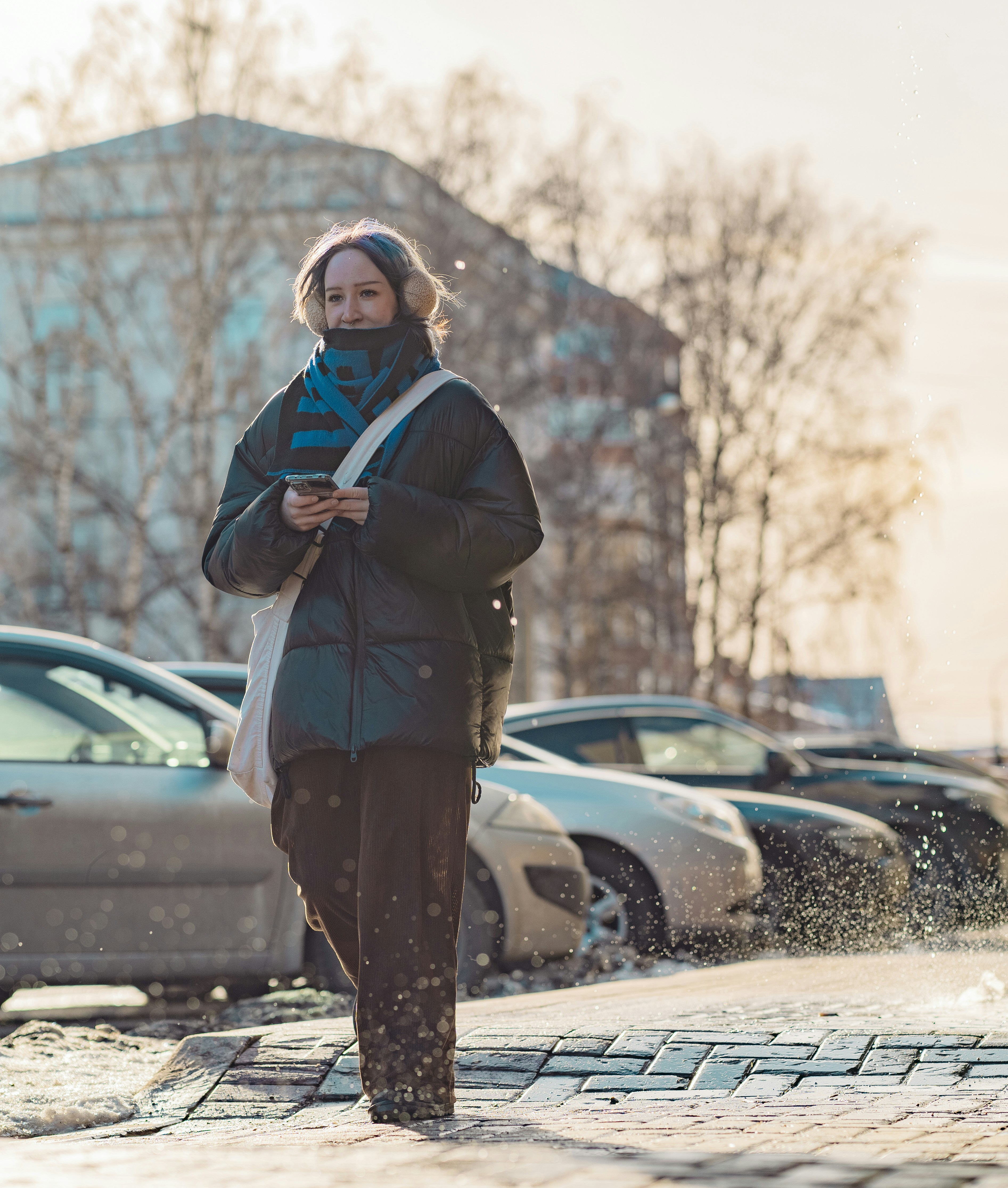 Backlit Pause: A scarfed pedestrian checks a phone beside sun-warmed parked cars