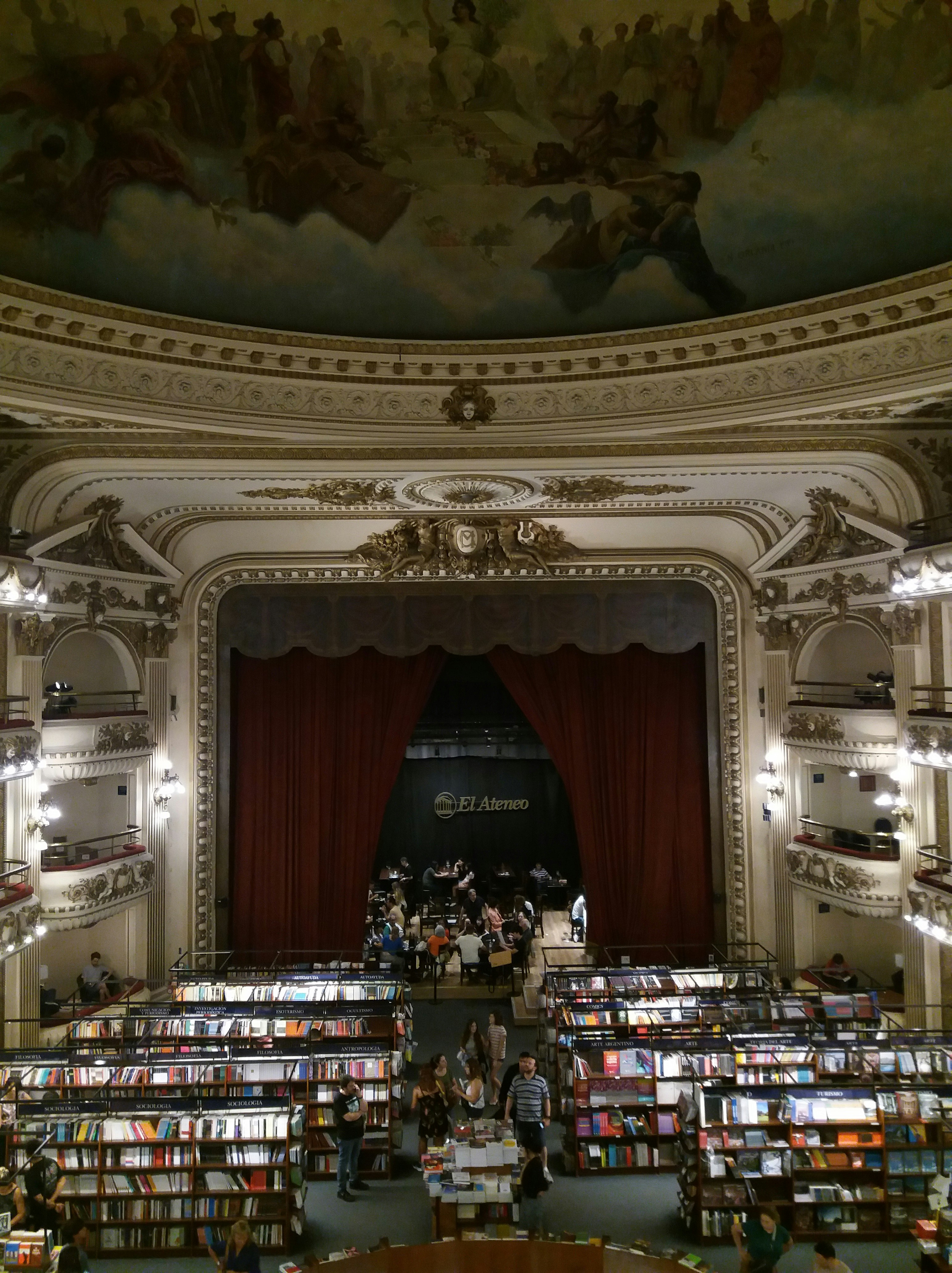 Ornate theater interior converted into a bookstore, with gilded arches, a painted ceiling, red curtains, and long shelves of books filling the stalls.