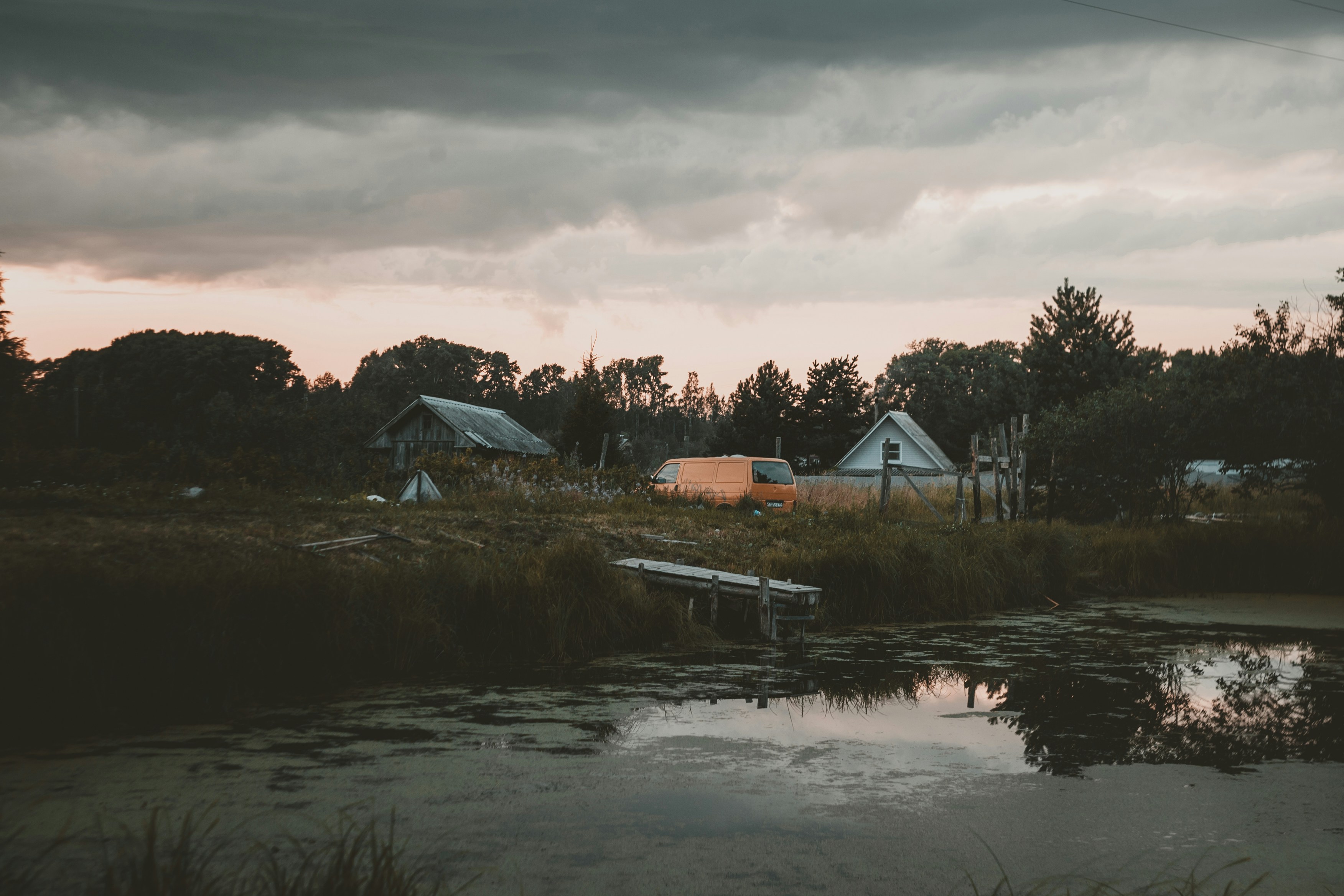 Abandoned buildings nestled beside a tranquil pond under a moody sky, evoking a sense of nostalgia and solitude.
