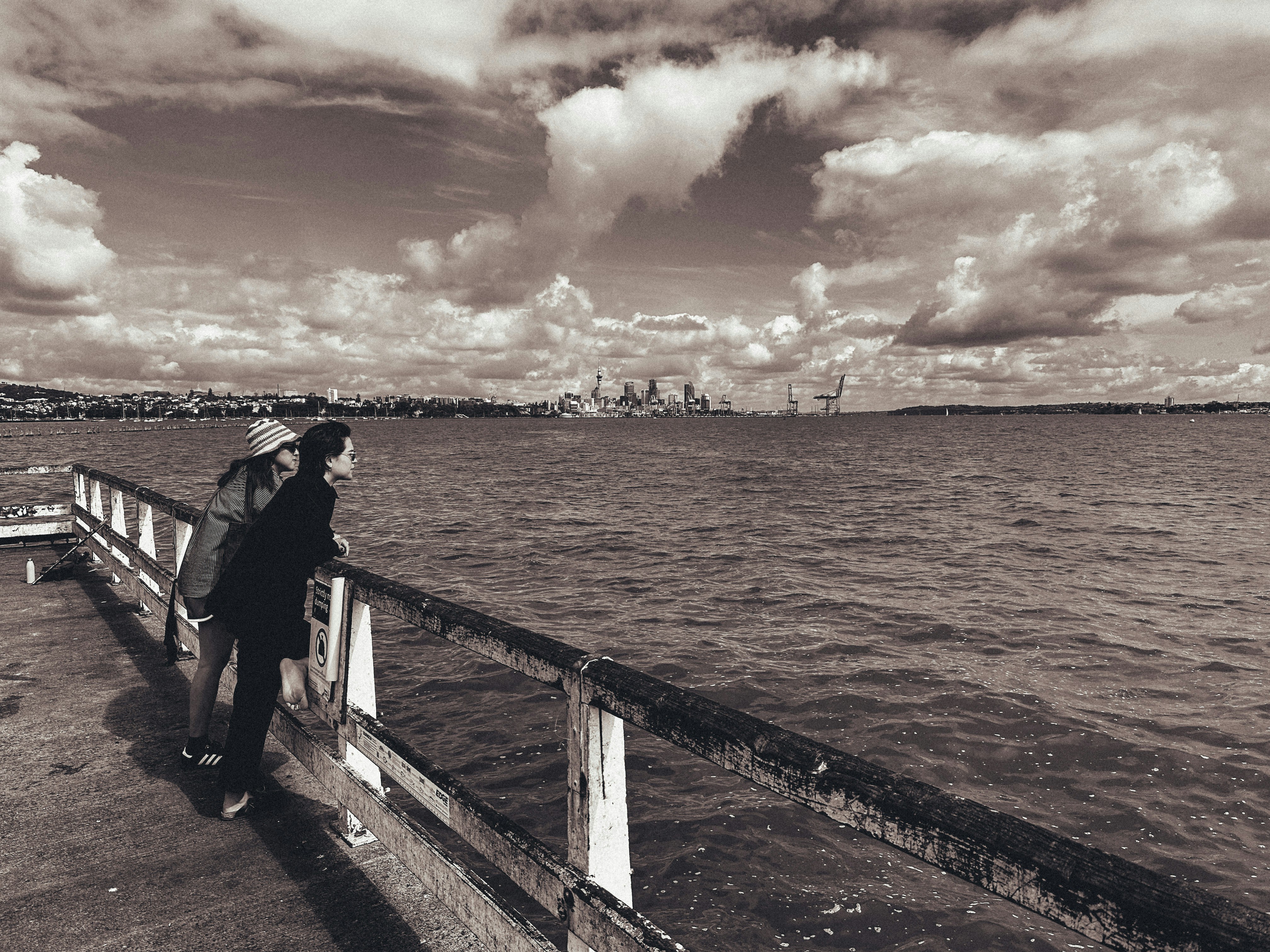 Sepia Harbor Moment: A couple at the railing gazes toward the distant city across the water