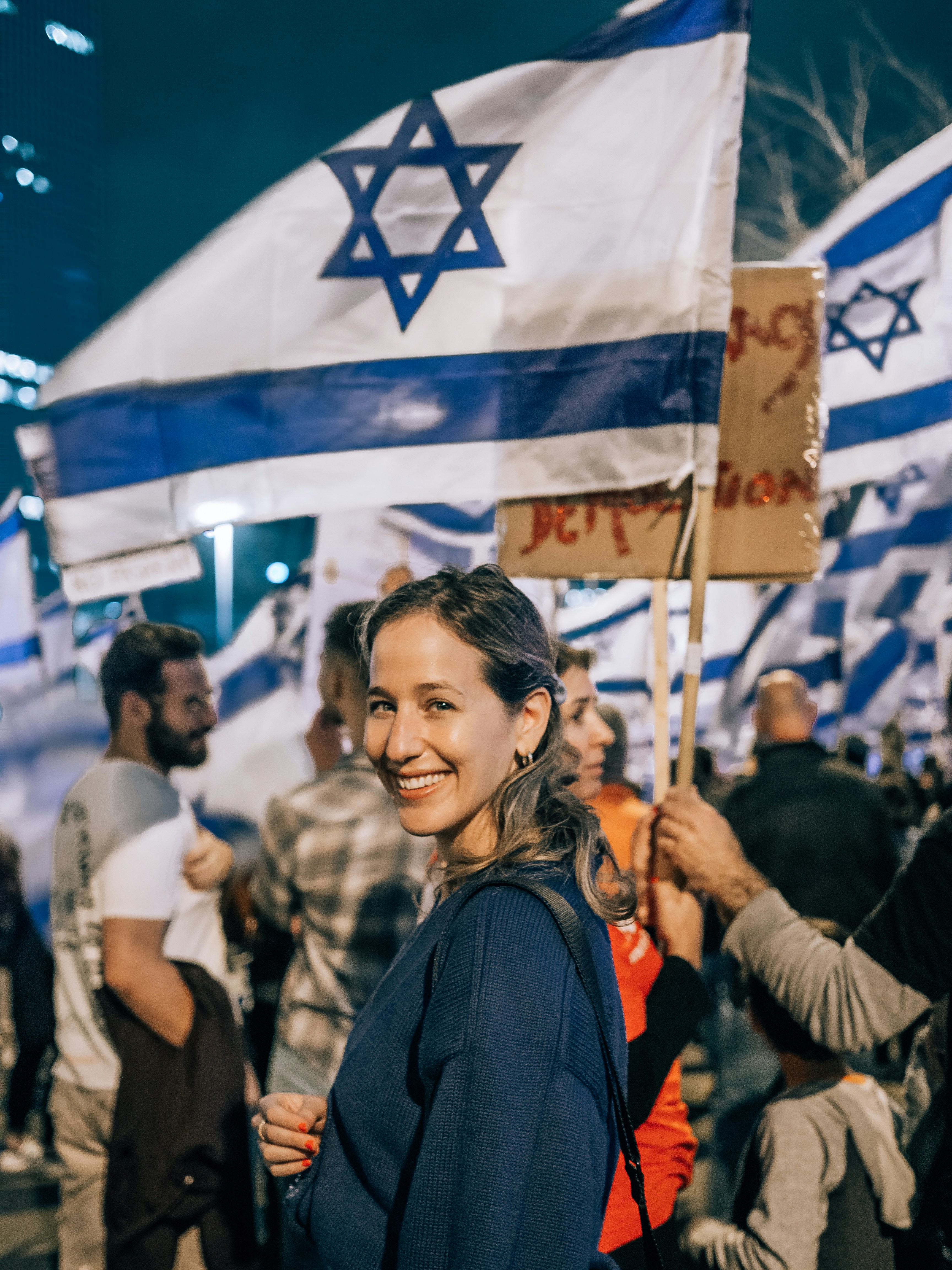 Woman smiling amidst a sea of waving Israeli flags at a vibrant gathering. The atmosphere reflects a strong sense of community and purpose.
