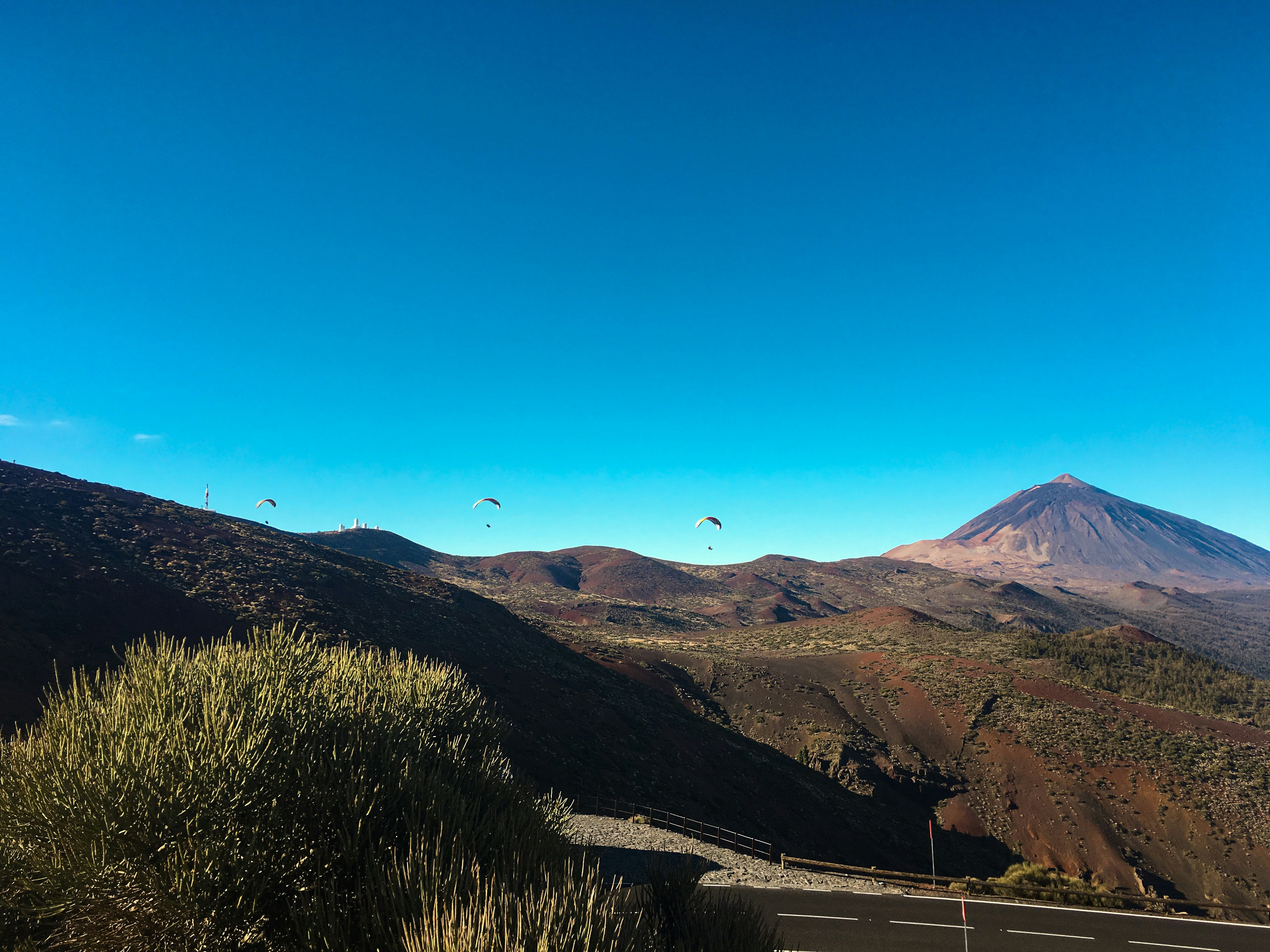 Volcanic ridge under a clear cobalt sky with several paragliders gliding above the hills; foreground features scrubby vegetation.