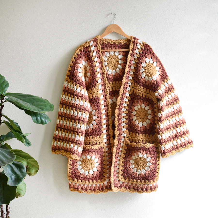 Handmade crocheted cardigan with large granny-square motifs in brown, cream, and mustard tones hanging on a wooden hanger against a white wall. A leafy plant frames the left side.