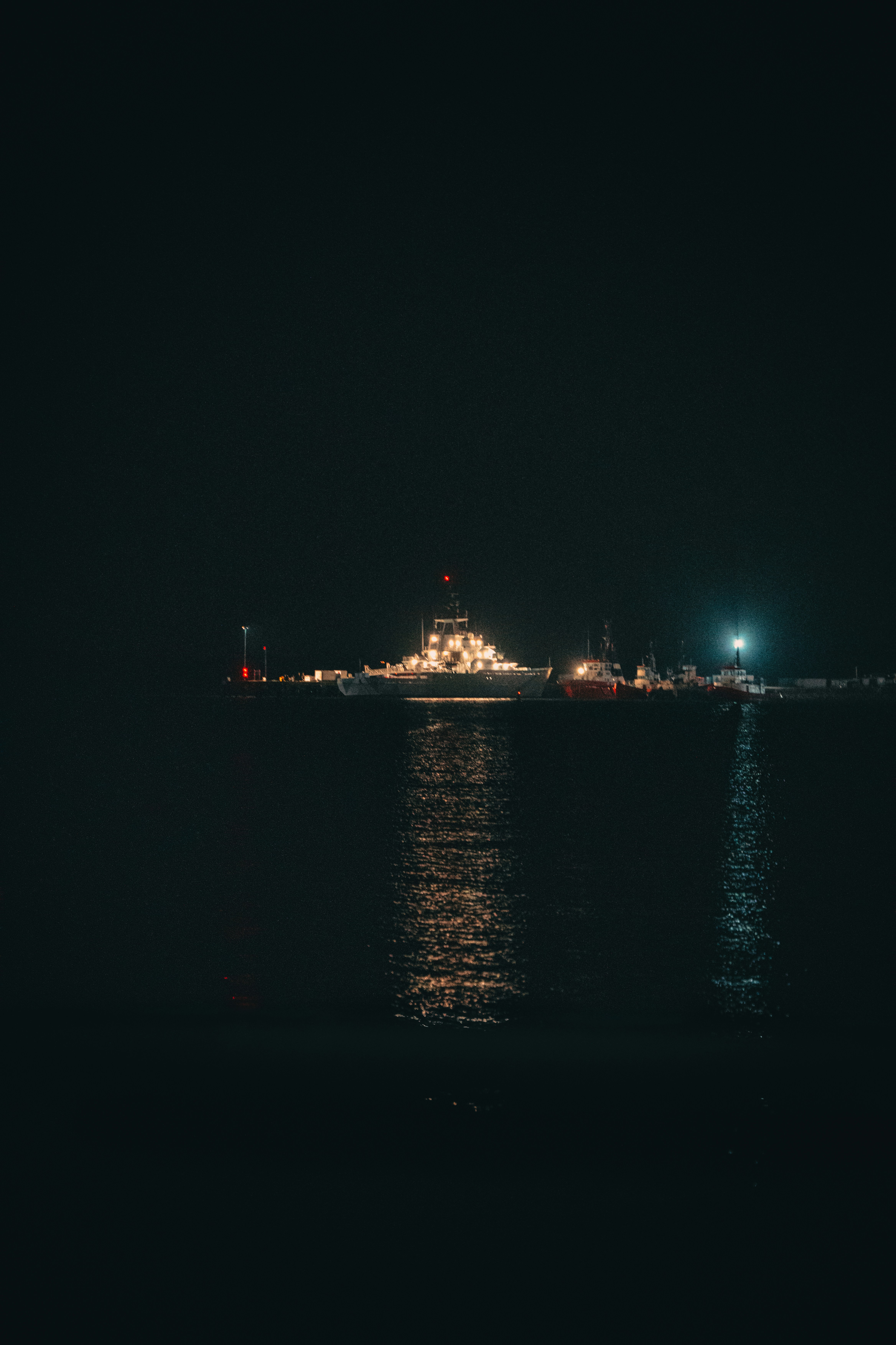 A brightly lit ship anchored in dark waters, reflecting its lights on the surface. The scene captures the tranquility of night maritime life.