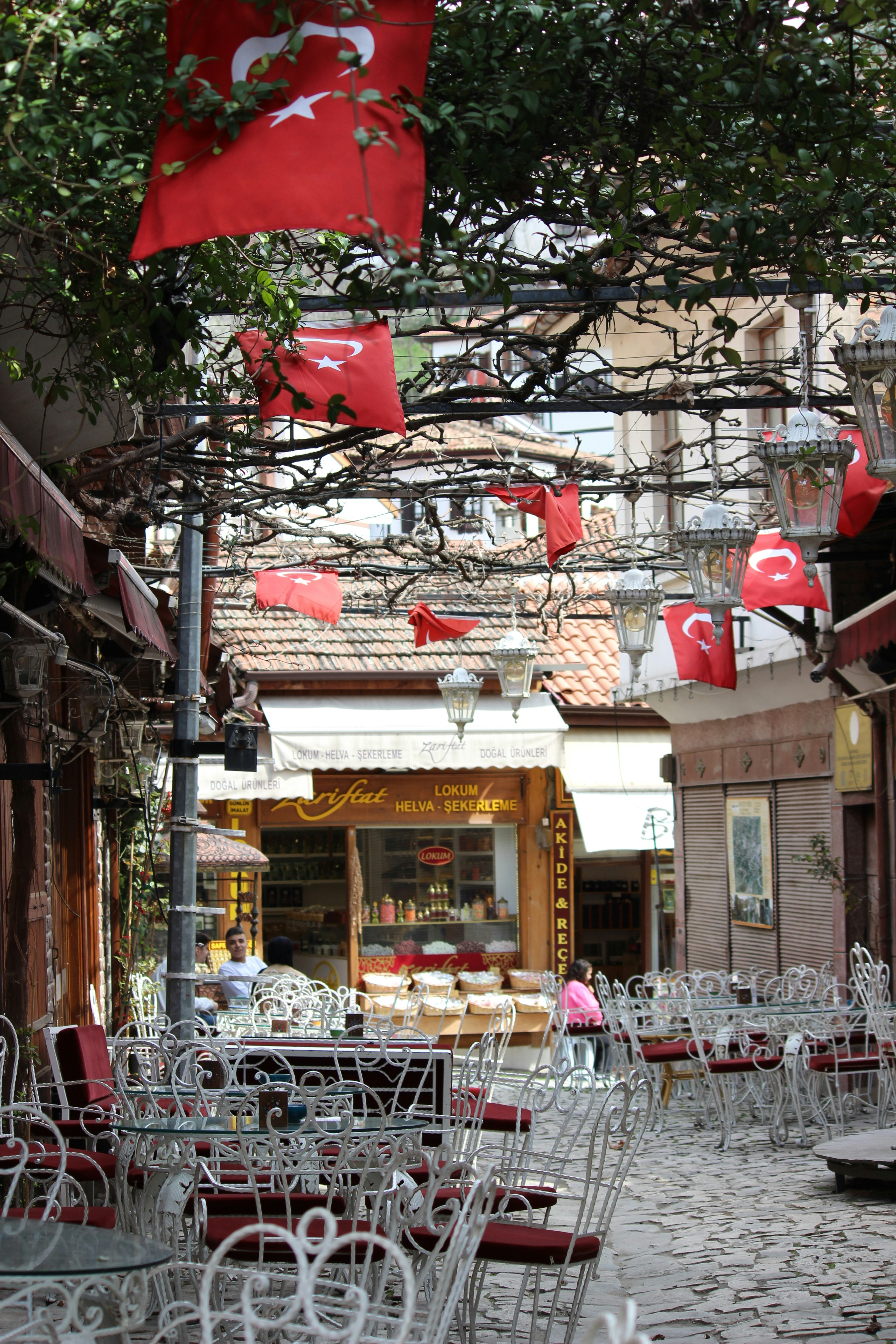 Quiet cobblestone alley with empty wrought iron café tables, adorned with red flags overhead.