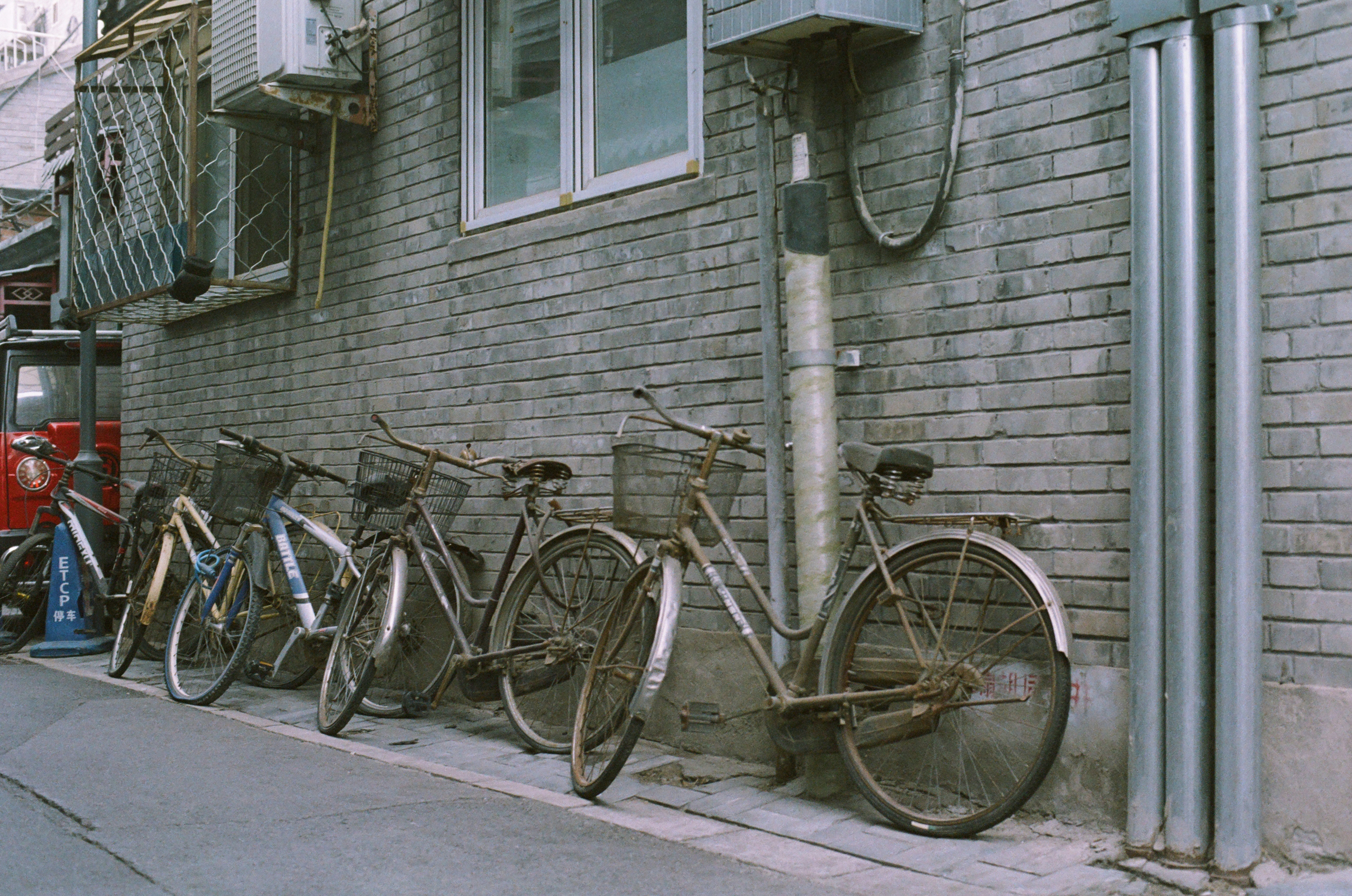 Row of weathered bicycles leaning against a brick wall along a narrow alley. The scene emphasizes urban decay and the quiet persistence of everyday objects.