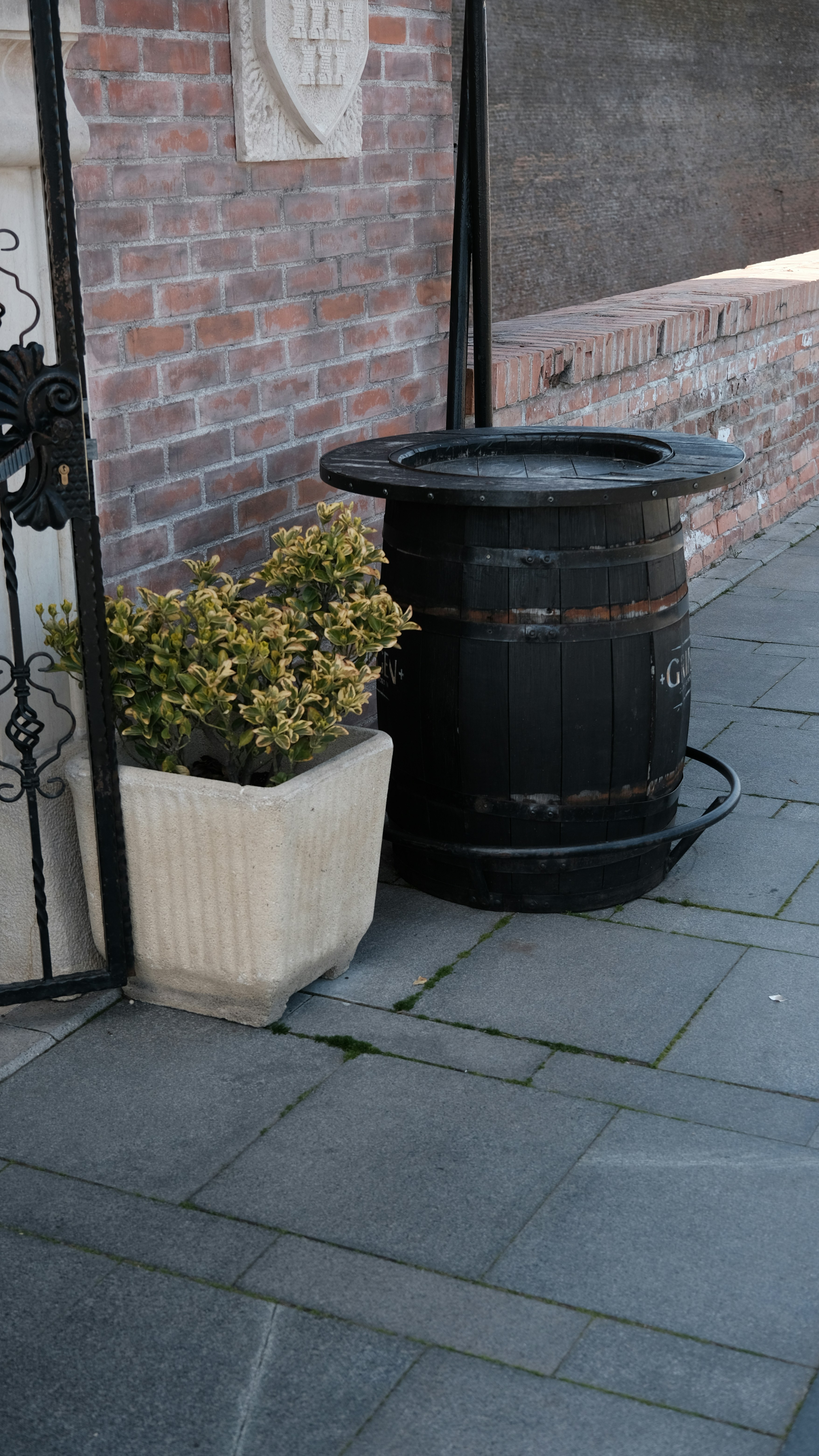 Black wooden barell and a green potted plant sitting near a brick wall.