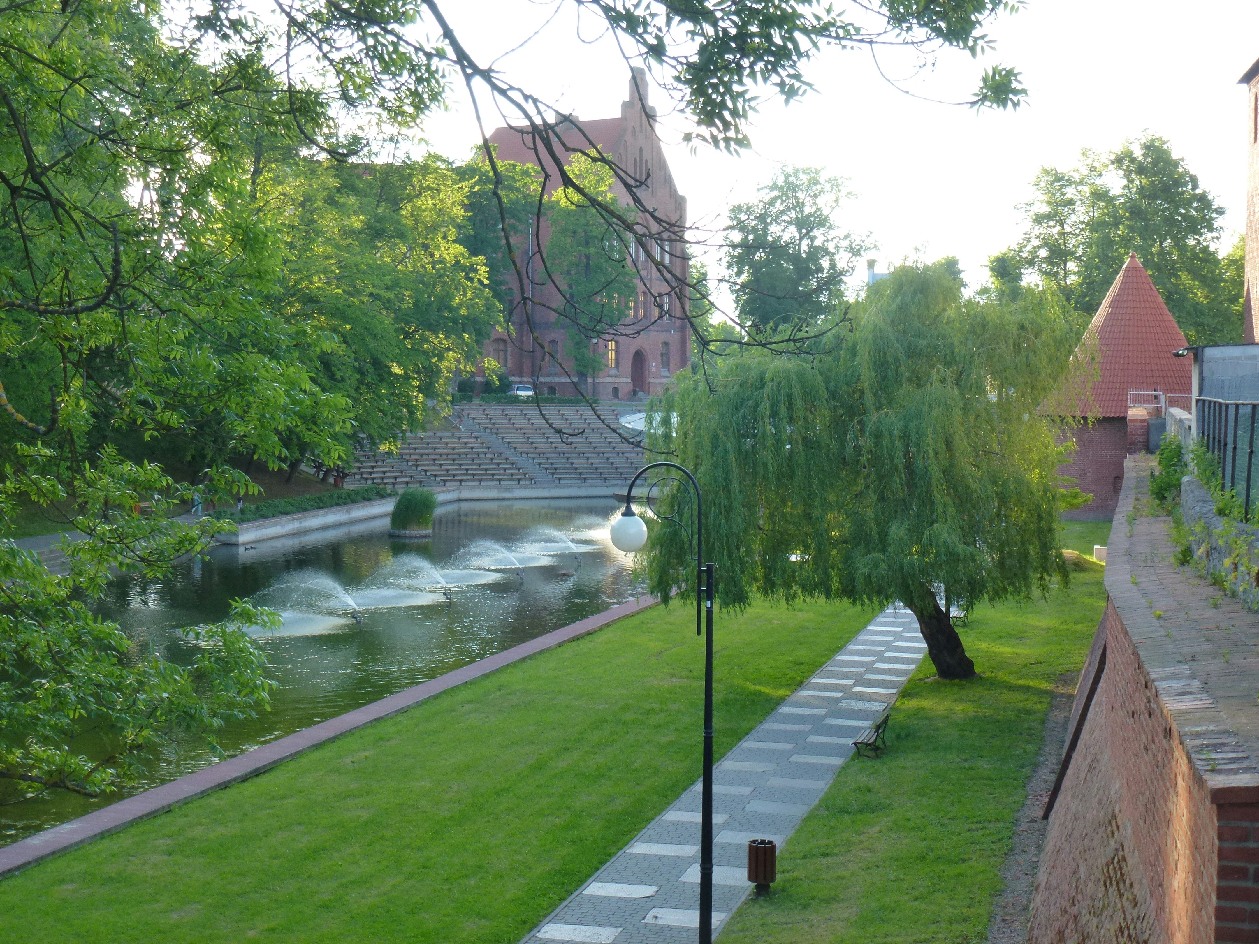 A tranquil riverside scene featuring lush greenery, a gentle fountain, and a pathway lined with trees. The historical architecture in the background adds depth to the landscape.