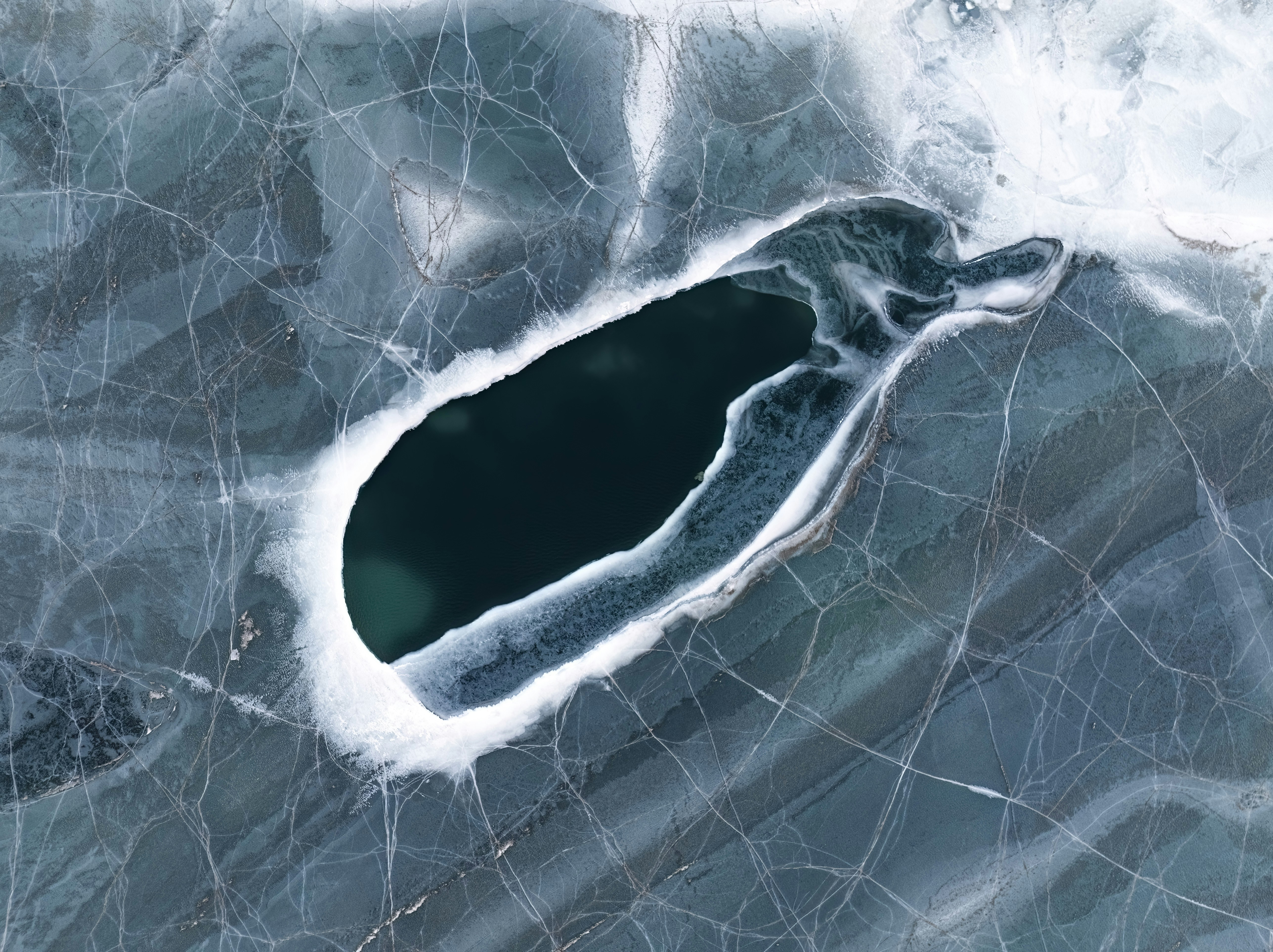 Dark water pool surrounded by intricate ice formations, revealing nature's delicate patterns. Aerial view showcases the stark contrast between the deep water and frozen surface.