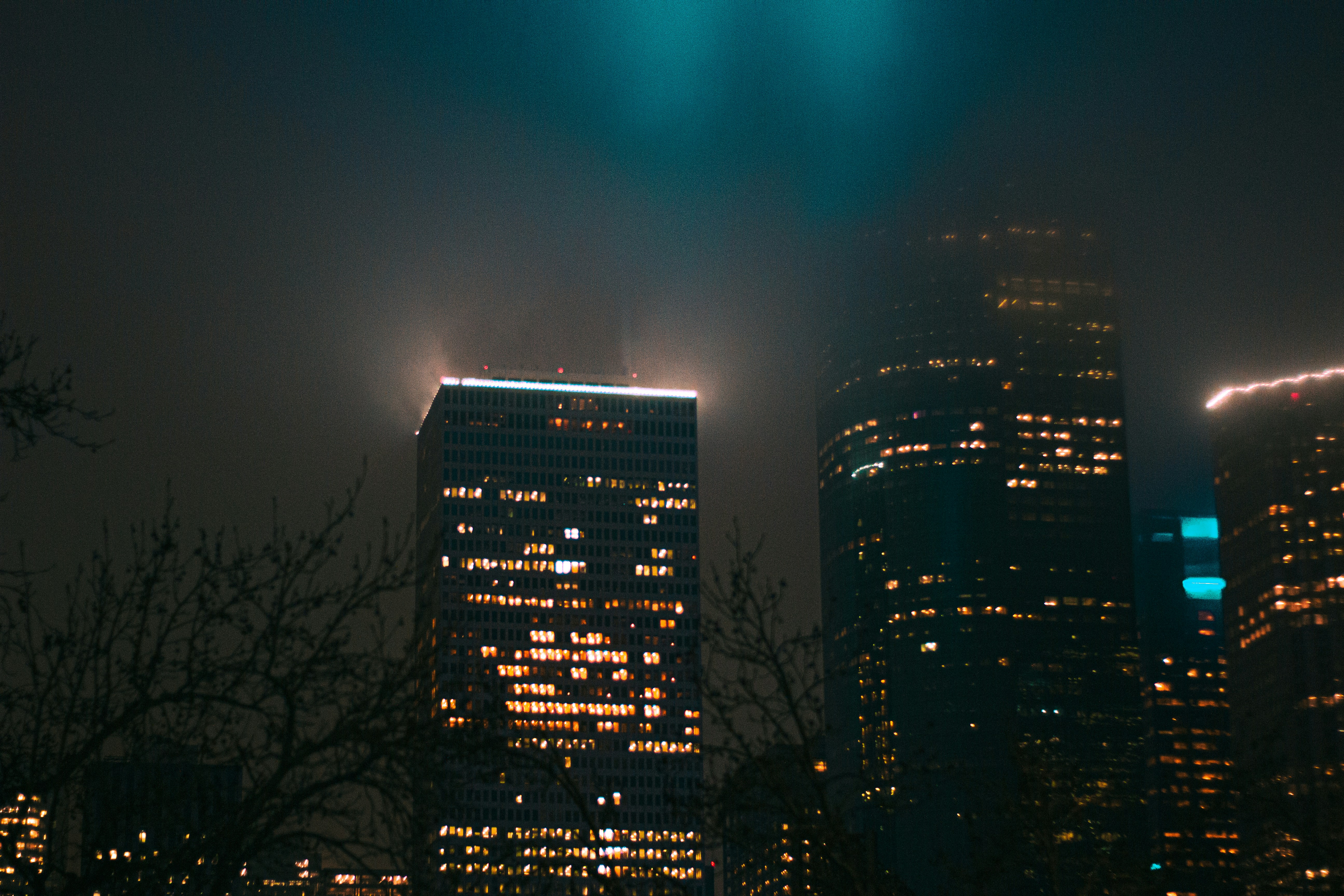 Illuminated skyscrapers rise through a veil of fog, showcasing a vibrant urban landscape at night.