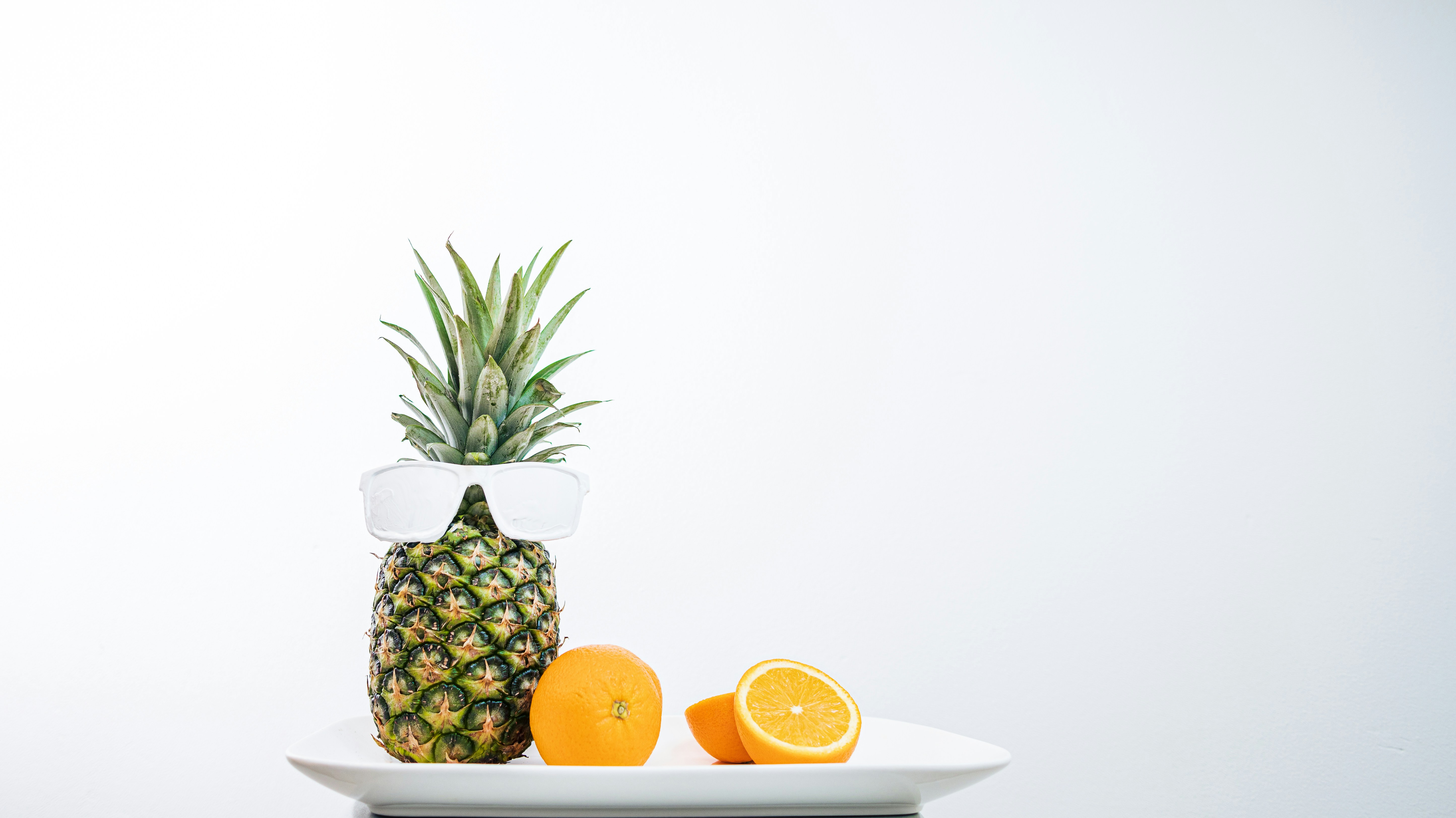 The picture depicts the joy of summer with a white plate placed on a white background. The plate holds two whole oranges, one of which is cut in half, and a whole pineapple wearing a pair of white sunglasses. The colors of the fruits along with the glasses give a vibrant and refreshing feel to the image, capturing the essence of summer. The picture represents the beauty of nature and the delicious taste of fresh fruits during the warm summer months.