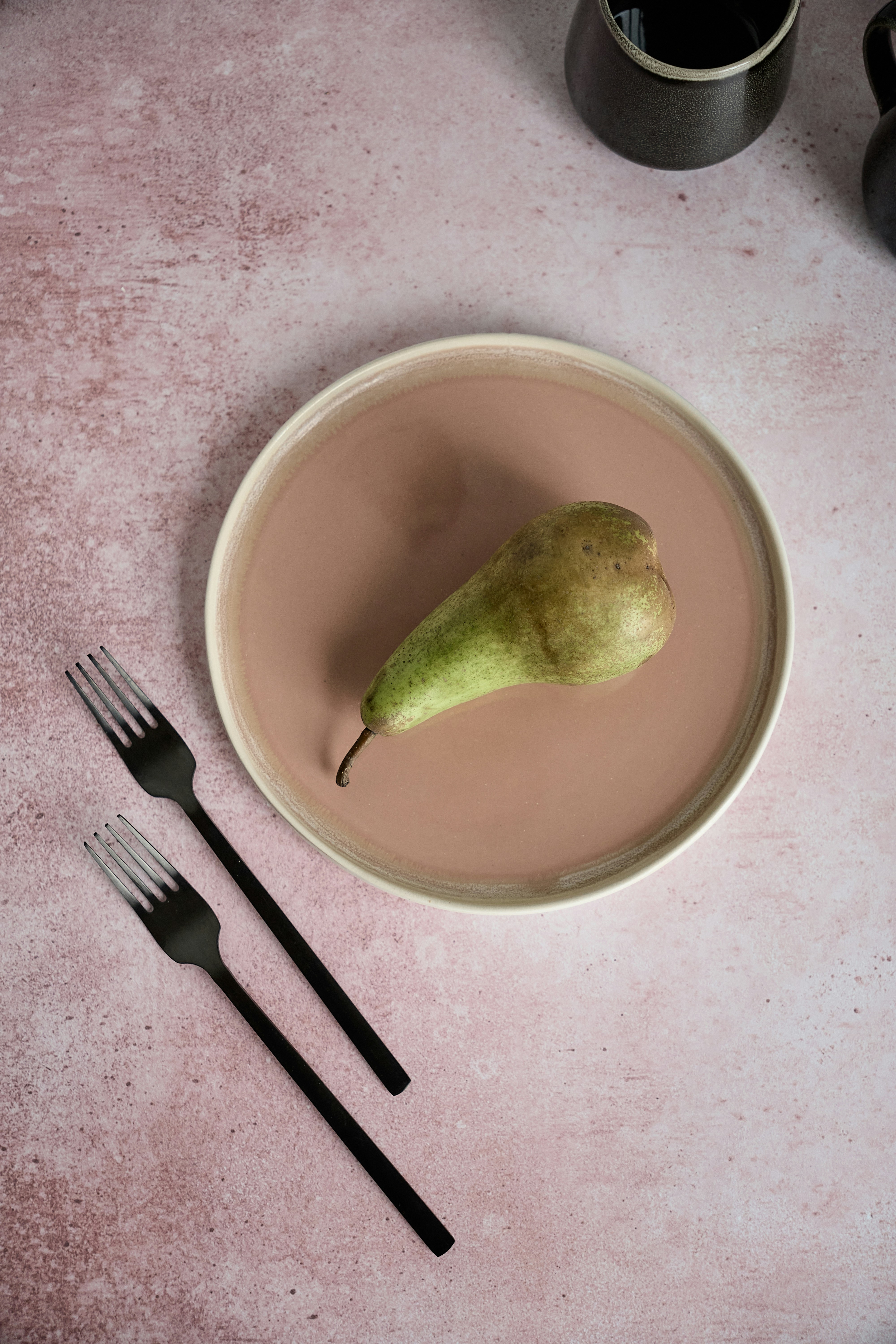 A green pear rests on a soft pink plate, accompanied by two black forks, creating a minimalist dining scene.