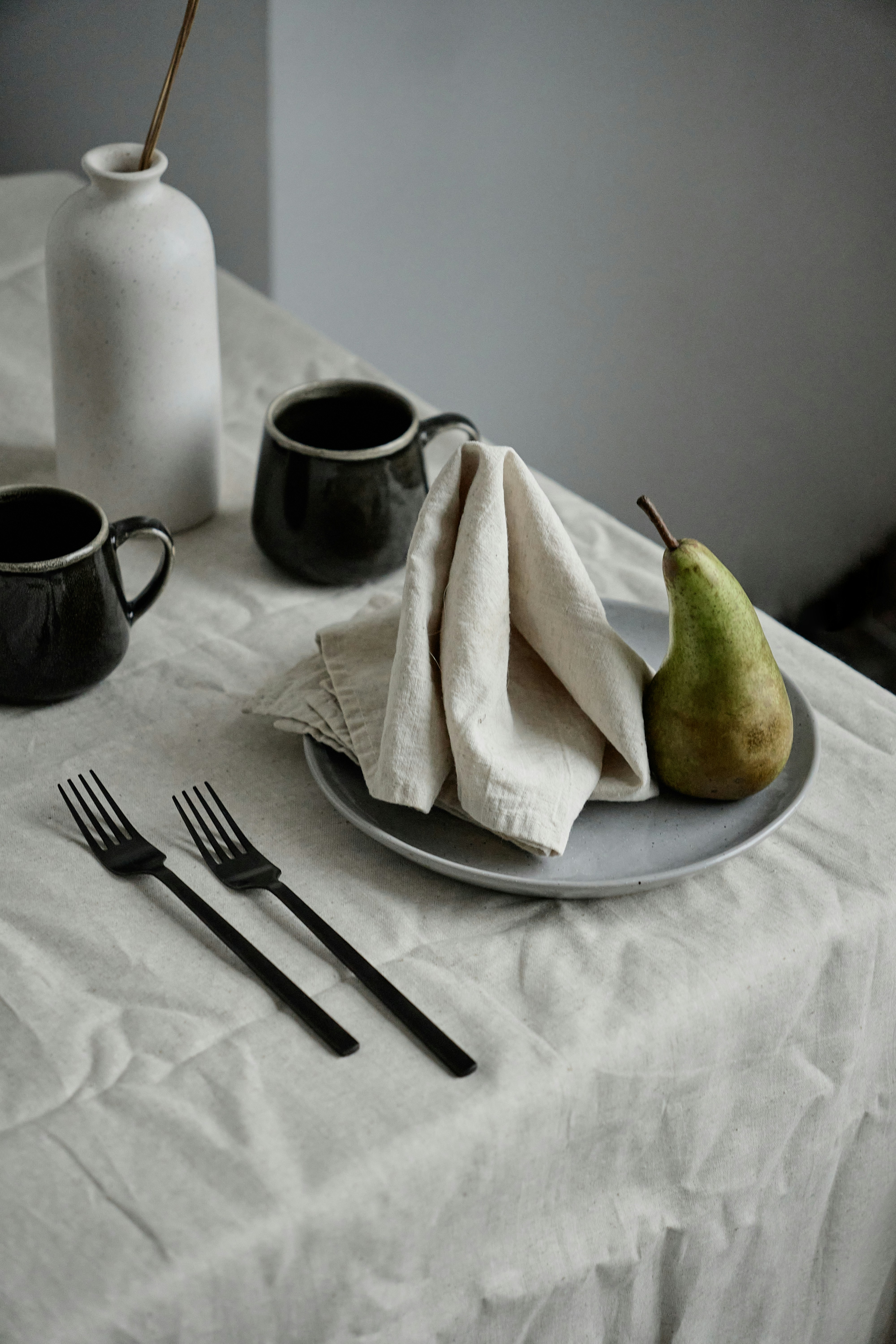 A quiet still-life photograph featuring a pear resting on a plate with a folded napkin, set among dark mugs and a ceramic bottle on a pale table.