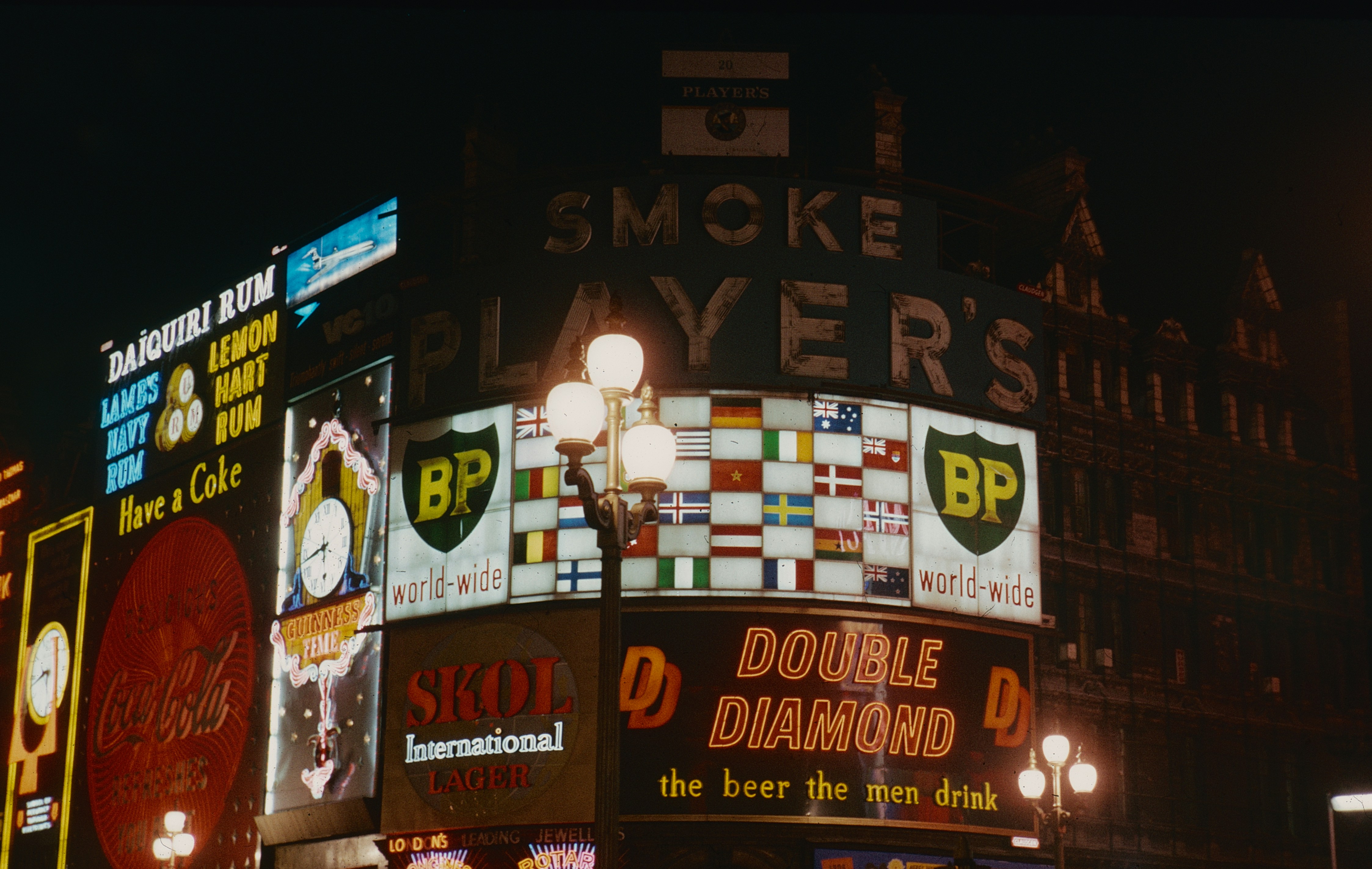 Vibrant billboards and neon signs illuminate a bustling urban corner at night, showcasing various brands and logos.