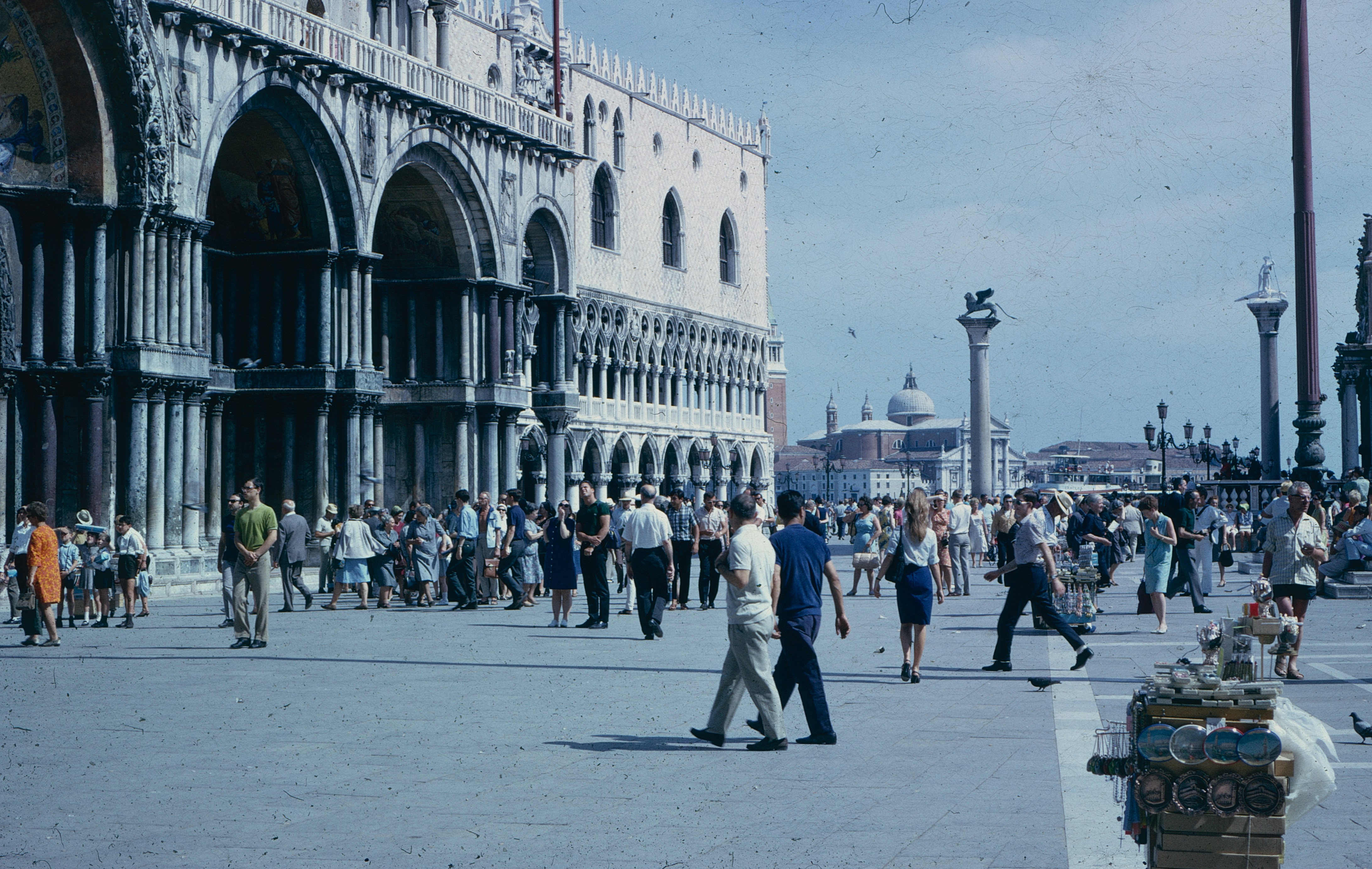 A photograph of a crowded plaza in front of an arched, ornate building. A couple walks toward the camera while other visitors linger in the sunlit space.