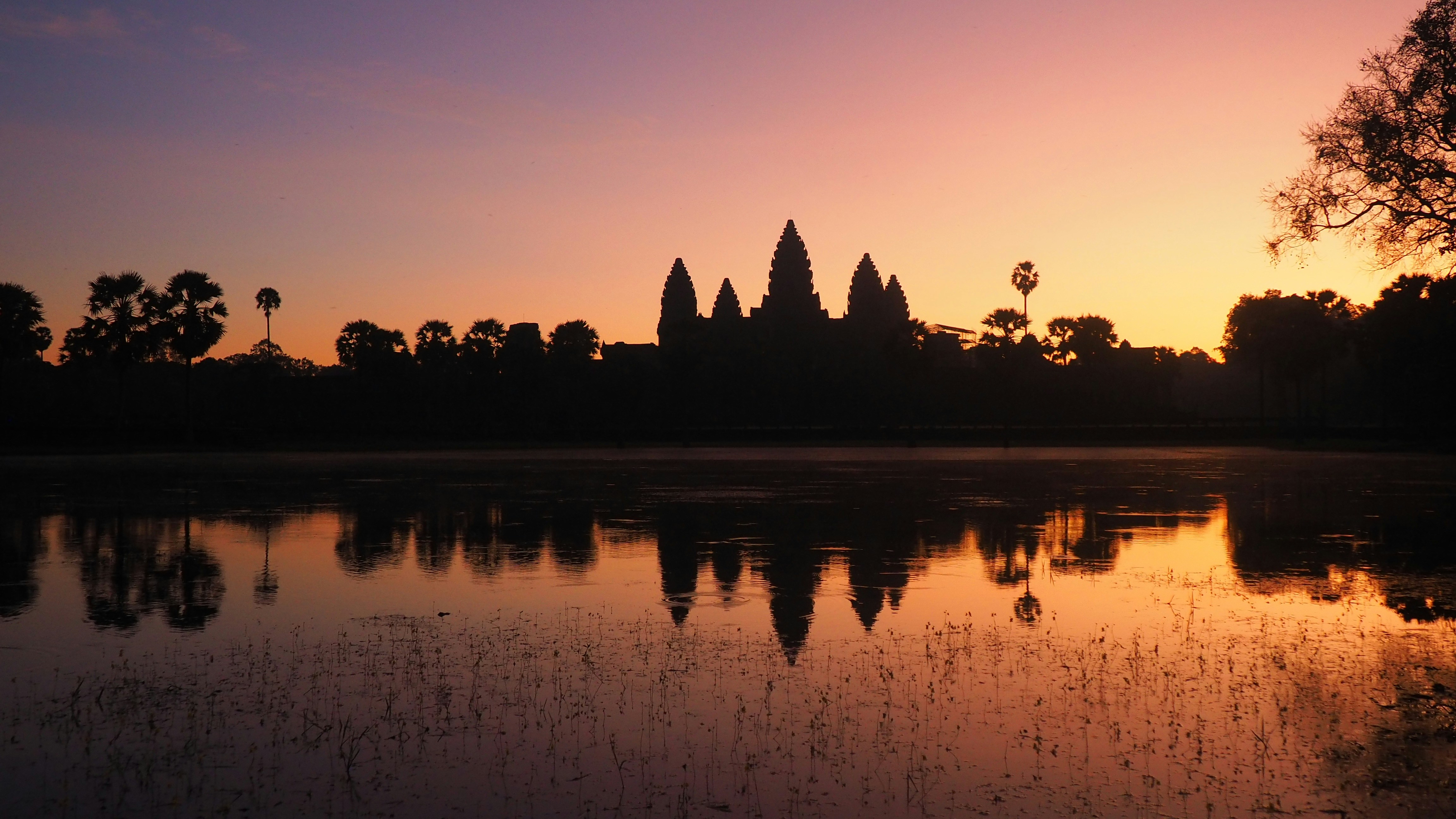 Angkor Wat silhouetted against a vibrant sunset, reflected in tranquil waters, showcasing the grandeur of ancient architecture.
