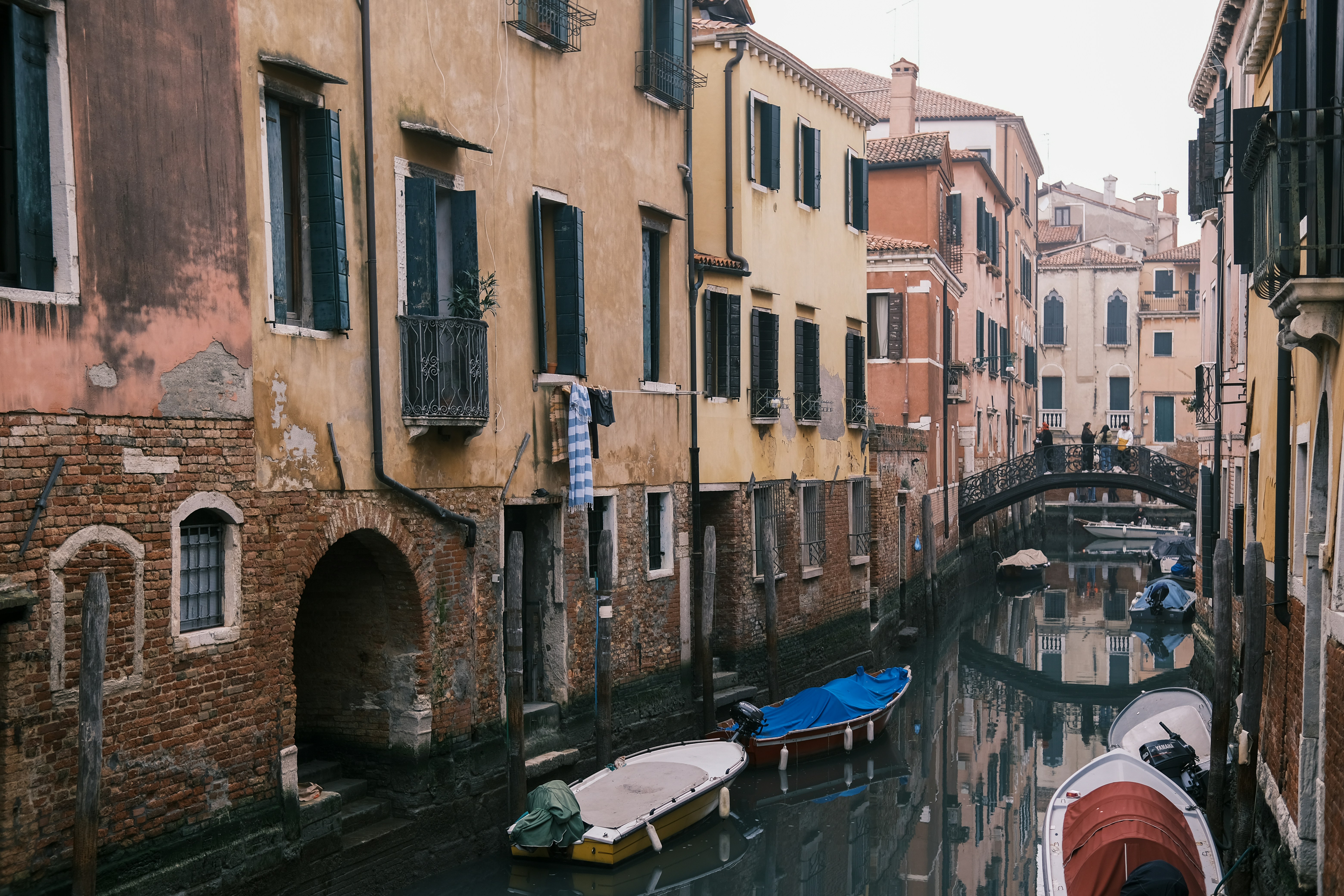 Narrow canal lined with rustic buildings and moored boats beneath a small bridge.