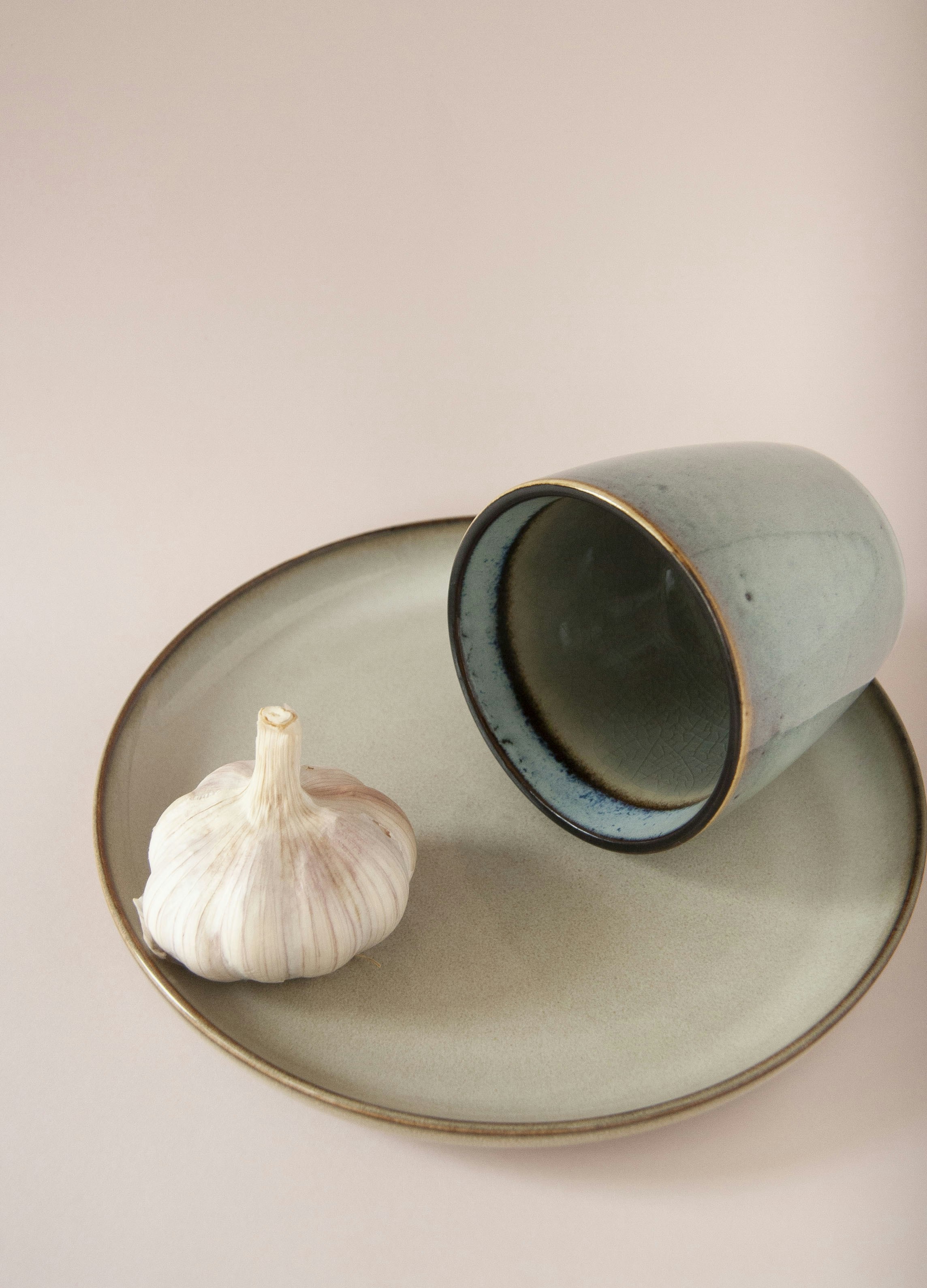 A soft-toned still-life photograph featuring a pale ceramic plate, a tilted teal mug, and a garlic bulb.