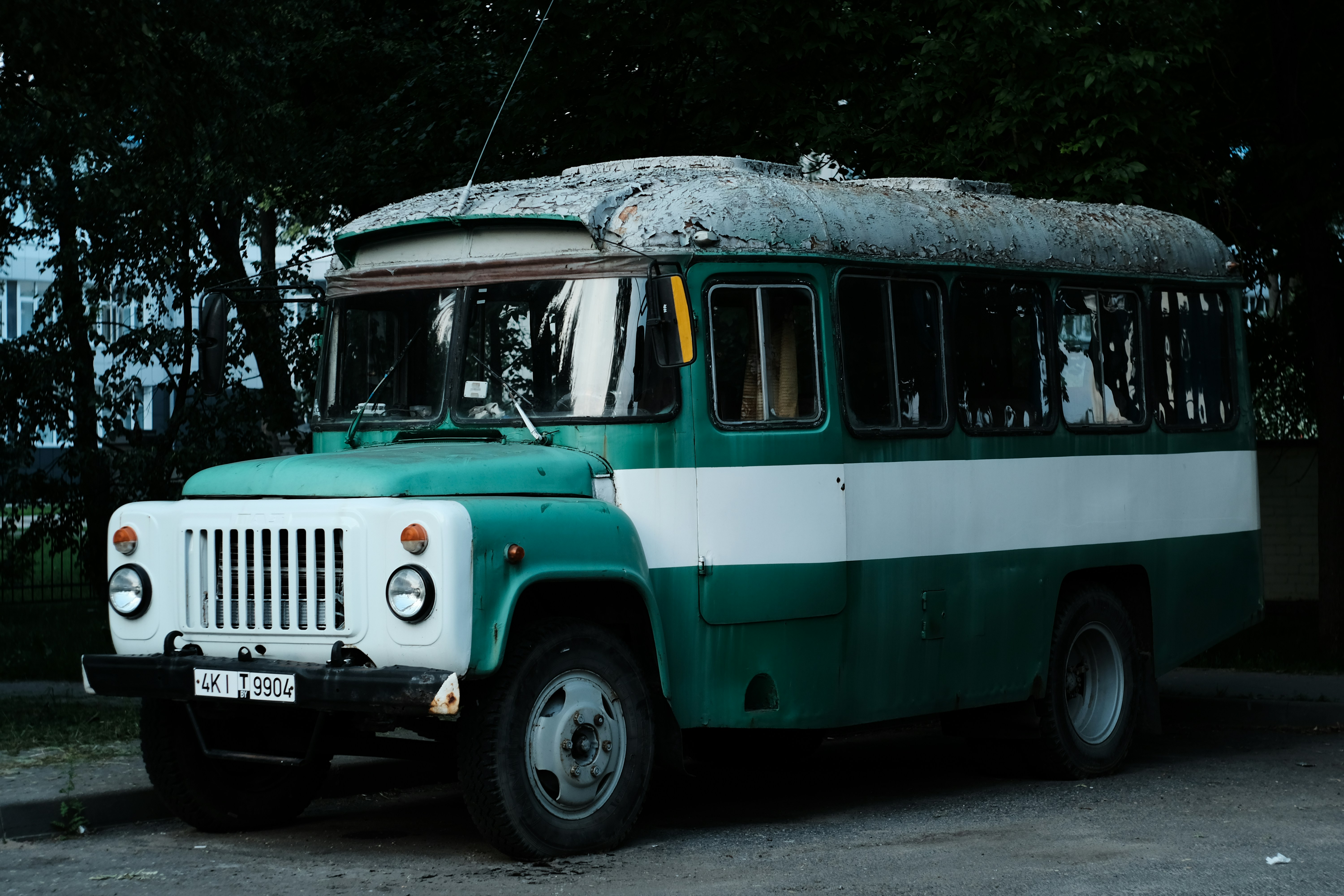 A weathered green and white bus parked under dim evening light with trees in the background.