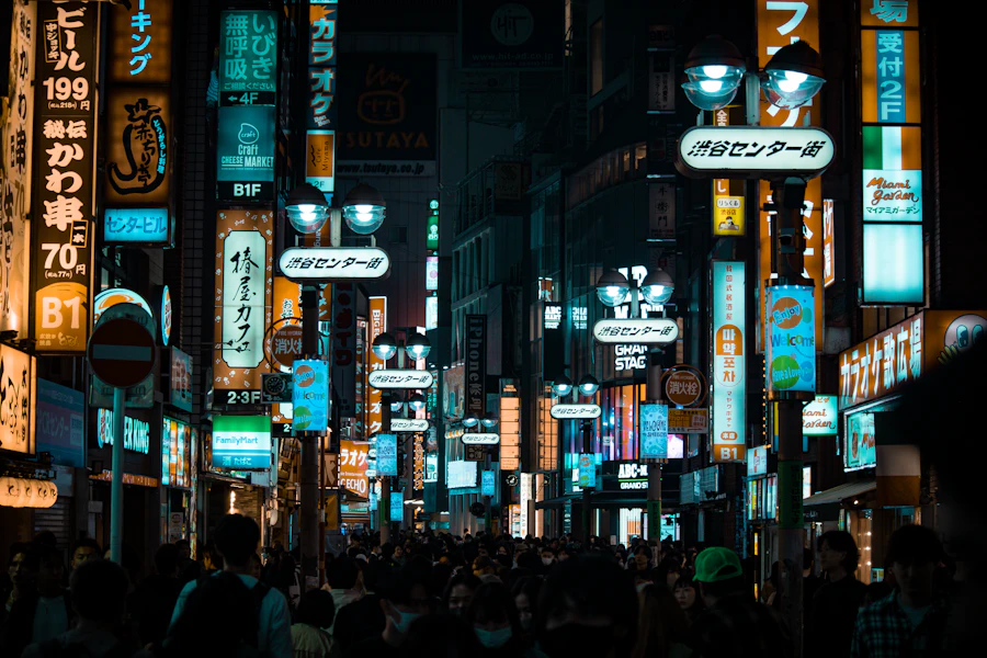 Crowded city street at night lined with glowing Japanese shop signs and blue street lamps. Dark silhouettes of pedestrians fill the foreground beneath the bright vertical signage.