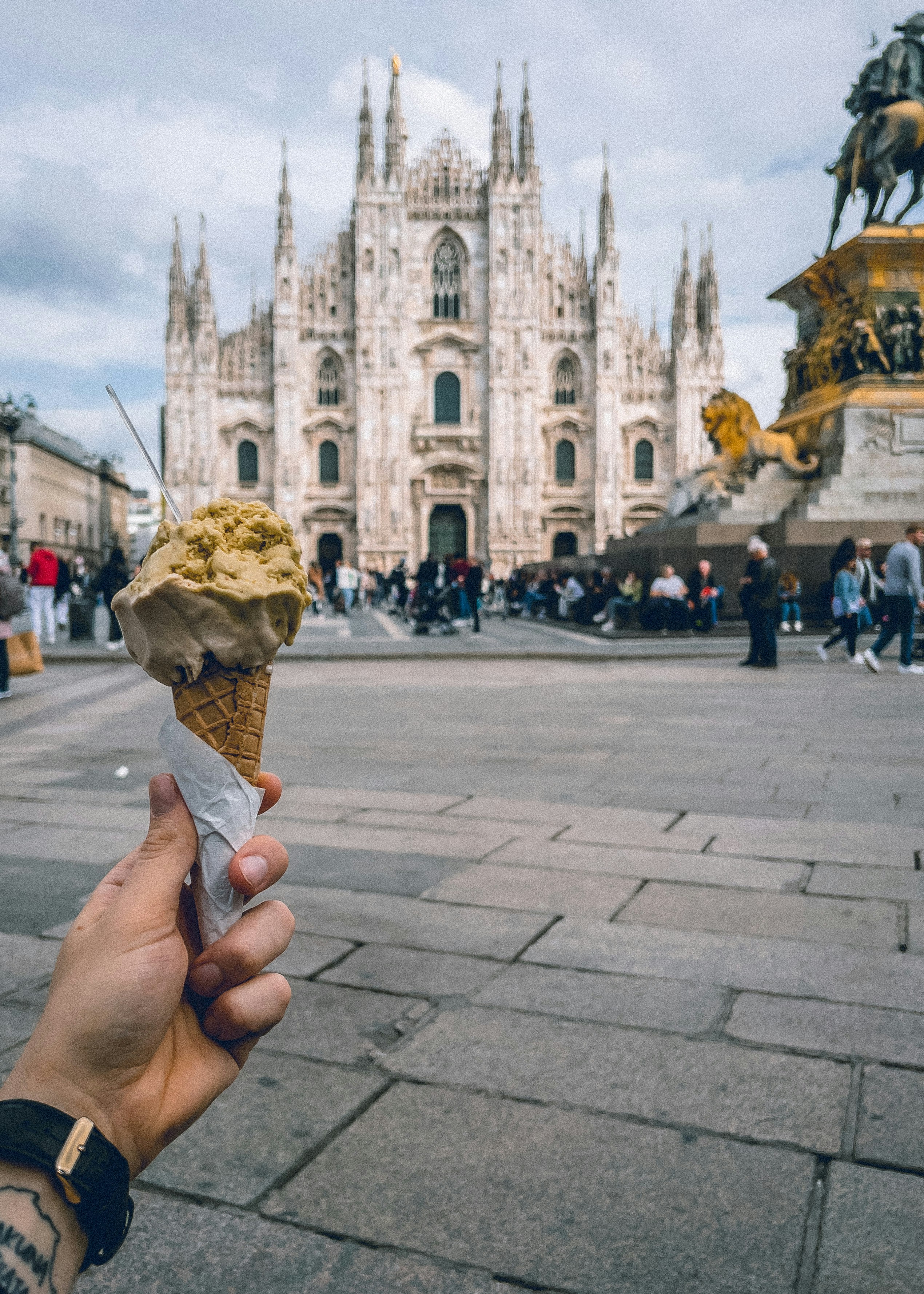 Hand holding a cone of gelato in front of a grand Gothic cathedral bustling with visitors. The scene captures the essence of Italian culture and culinary delight.