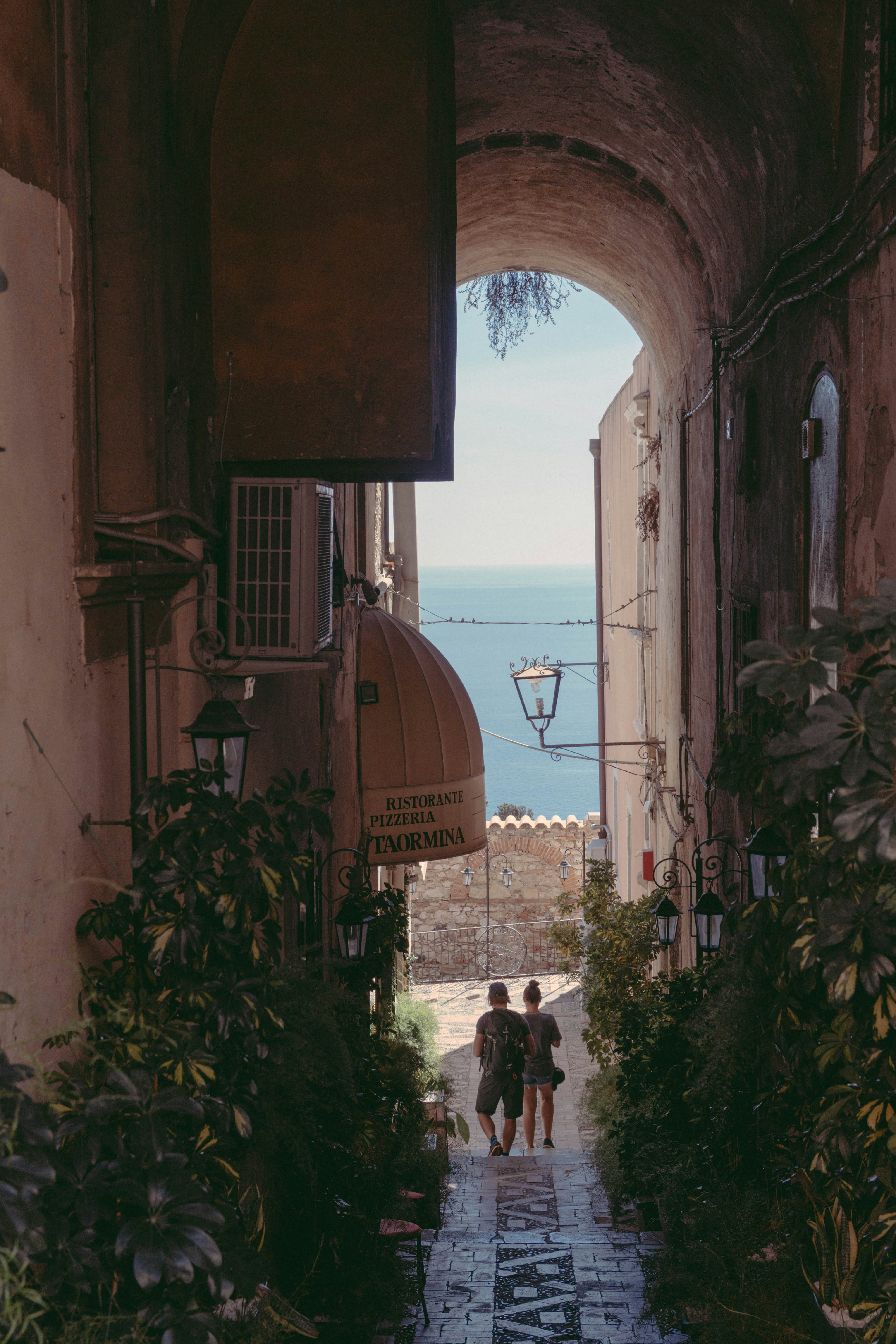 Couple walking hand in hand down a charming alleyway, framed by lush greenery and historic architecture, with a view of the sea in the distance.