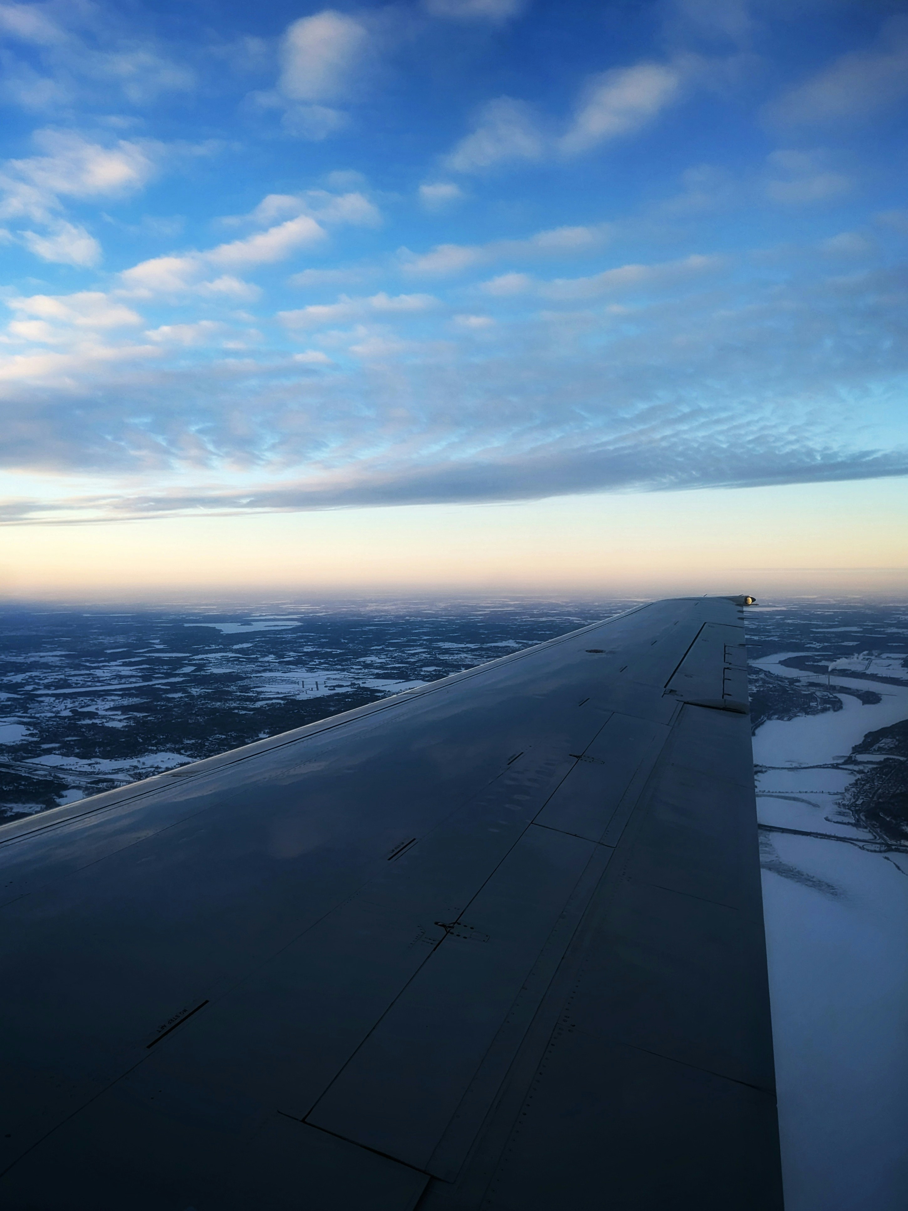 Airplane wing stretches toward a pale horizon as the setting sun bathes the clouds in warm hues.