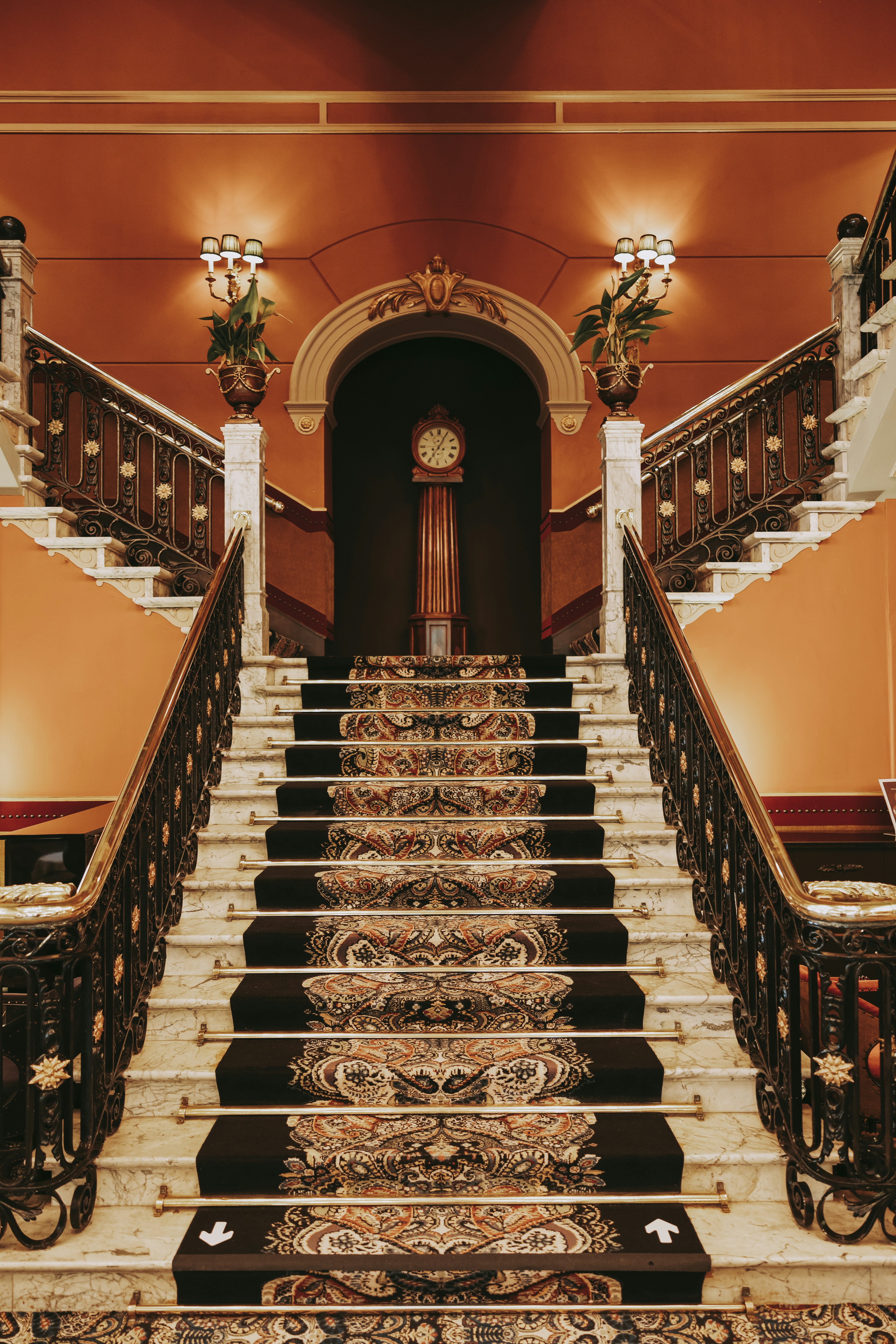 Intricately designed staircase adorned with a luxurious carpet, leading to an ornate clock and elegant decor. The warm tones enhance the opulence of the setting.