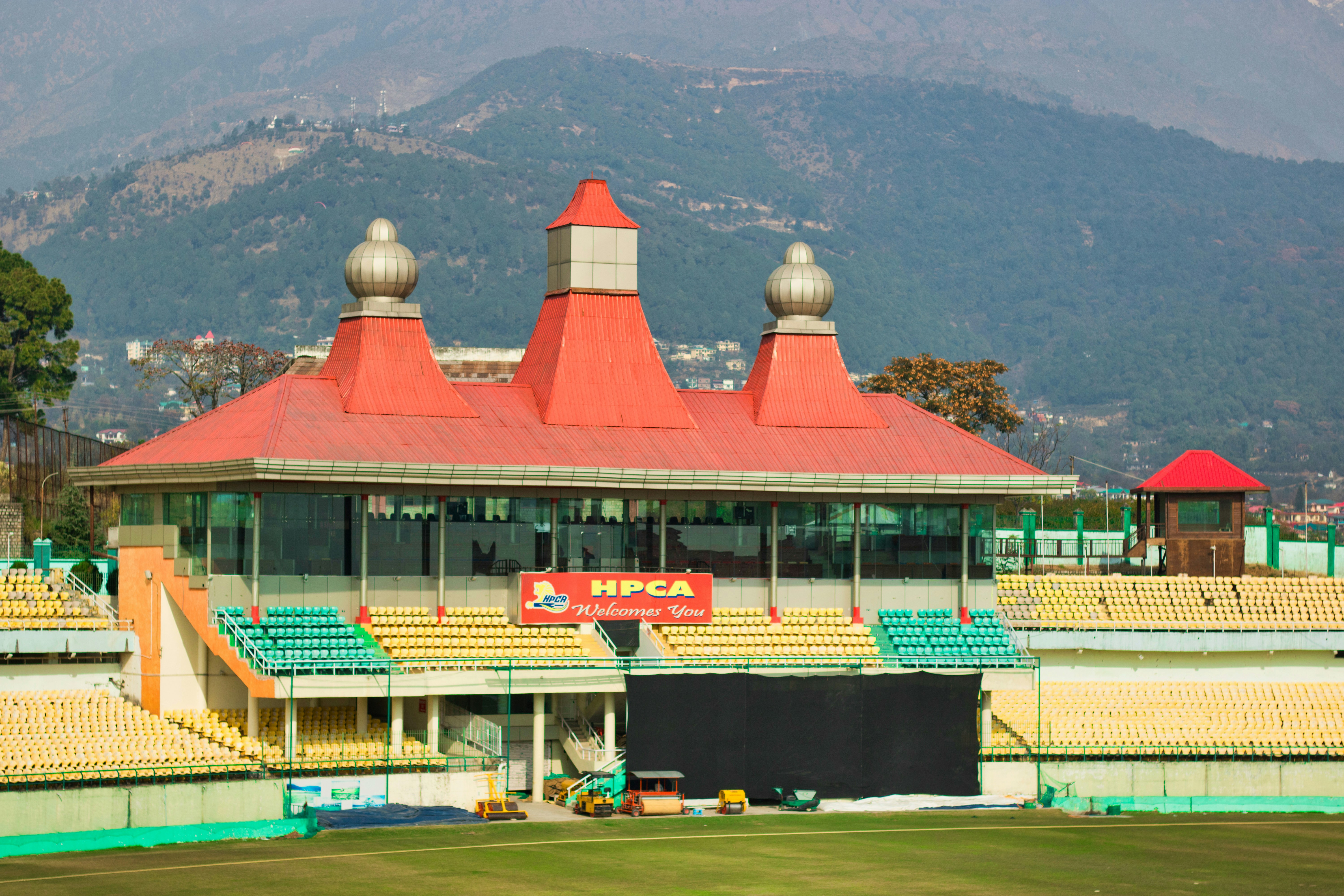 The HPCA Stadium showcases a unique architectural style with its striking red roof and modern glass facade, set against a backdrop of mountains.