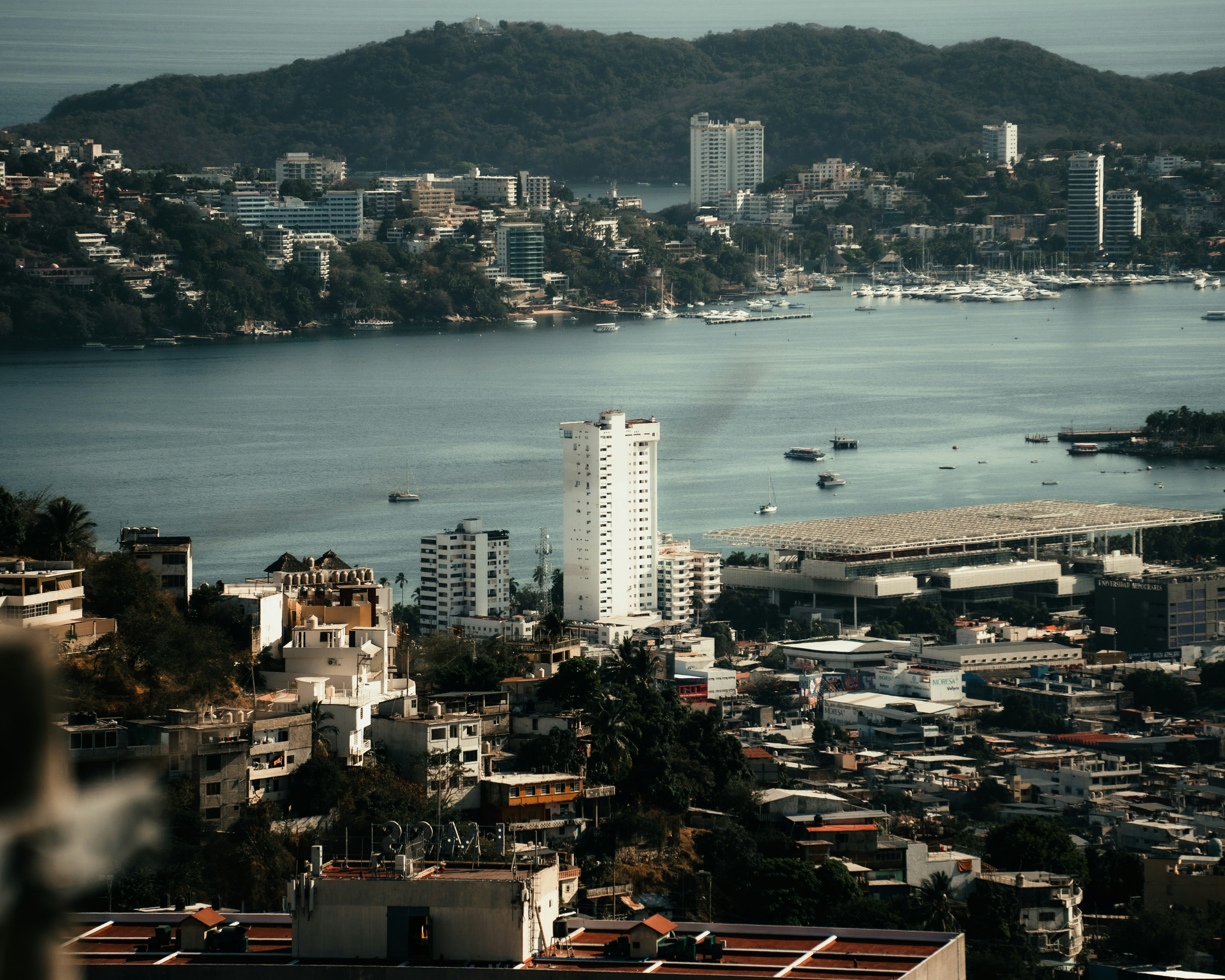 Hillside cityscape with buildings leading to a tranquil bay, framed by distant mountains.