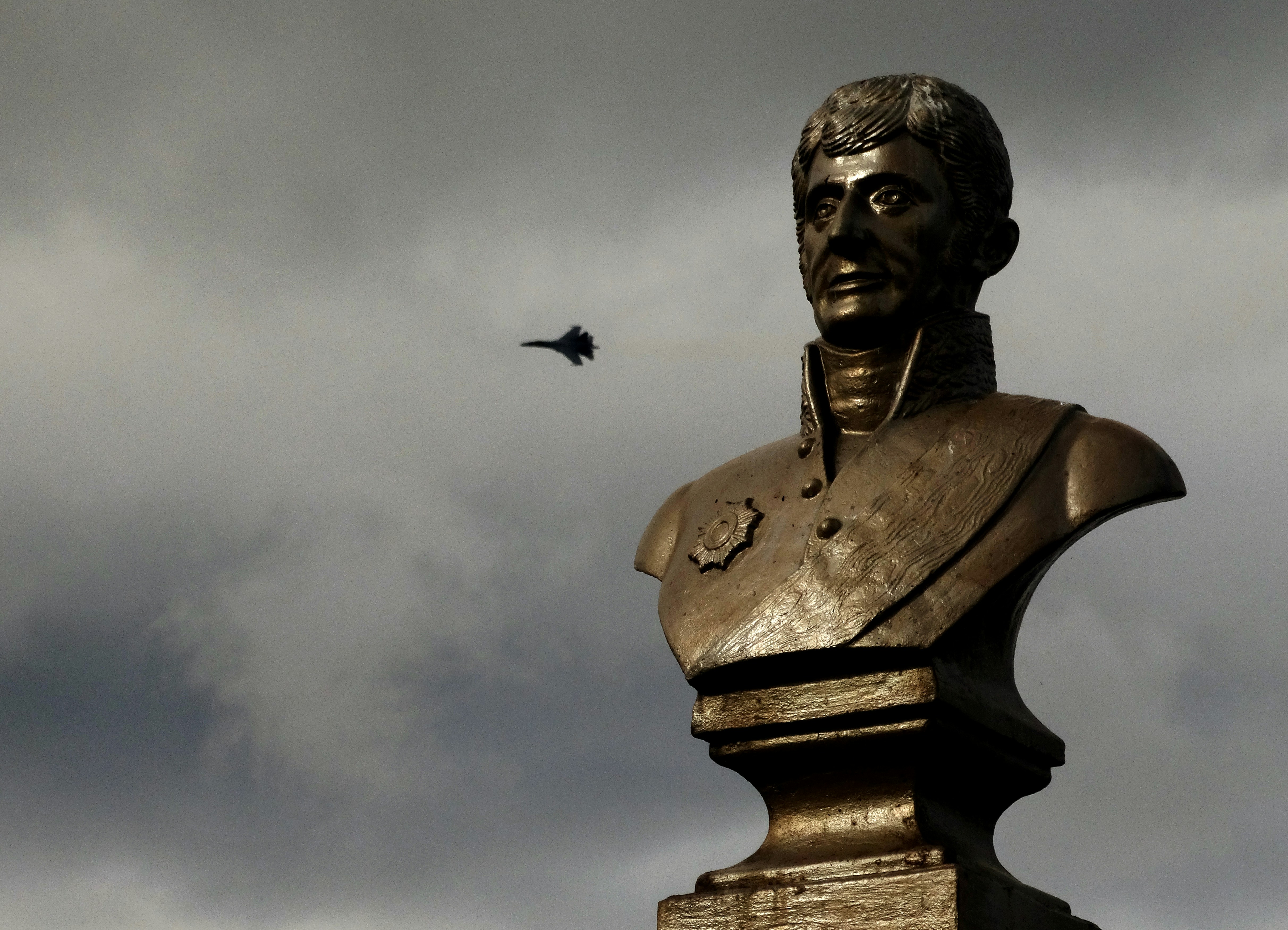 Bronze bust of a historical figure against a dramatic cloudy sky, with a silhouette of a bird flying in the background.