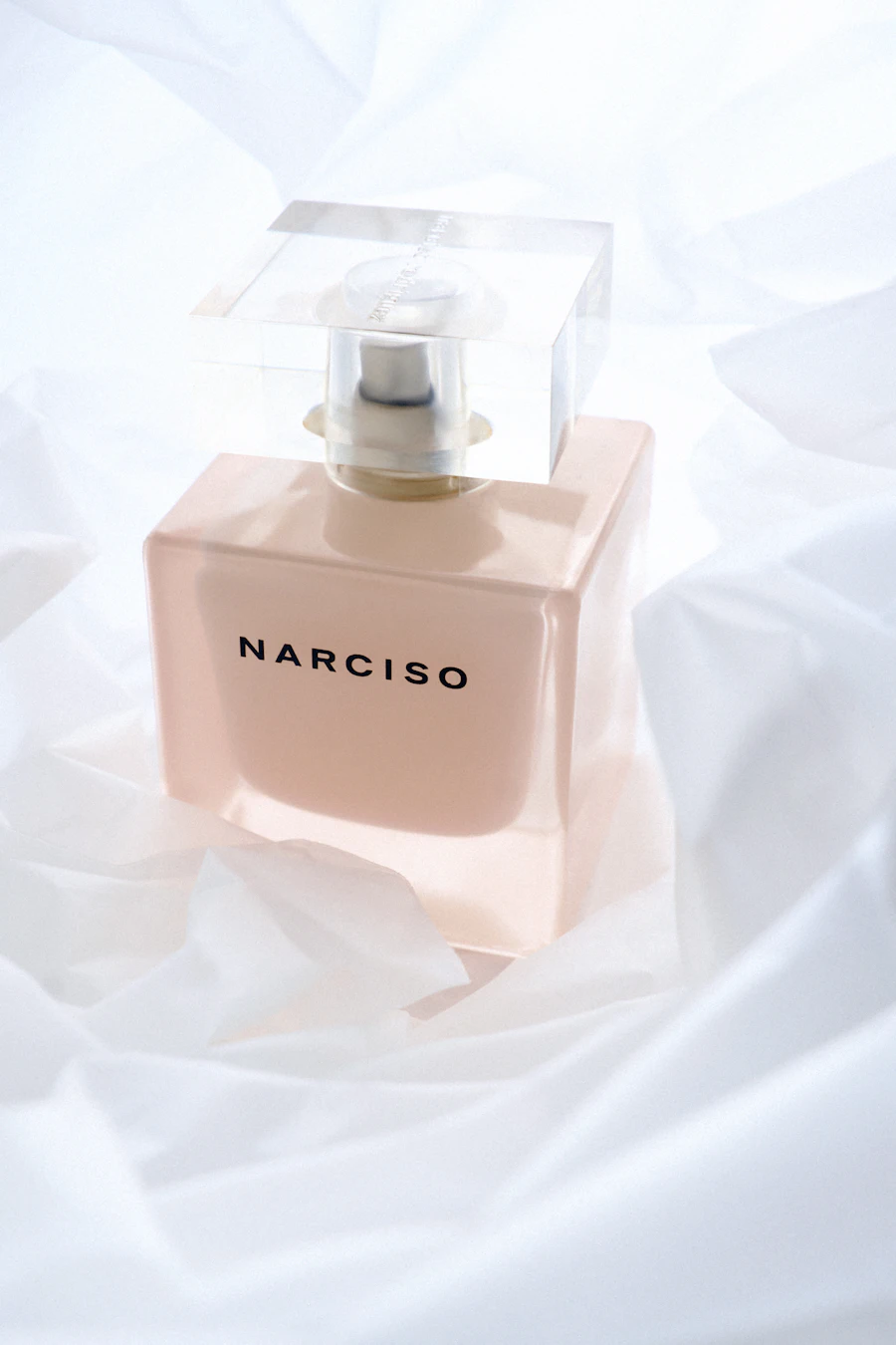 Pale pink Narciso perfume bottle with a clear square cap nestled in airy white fabric. Bright, dreamy lighting gives the product a clean, glossy look.