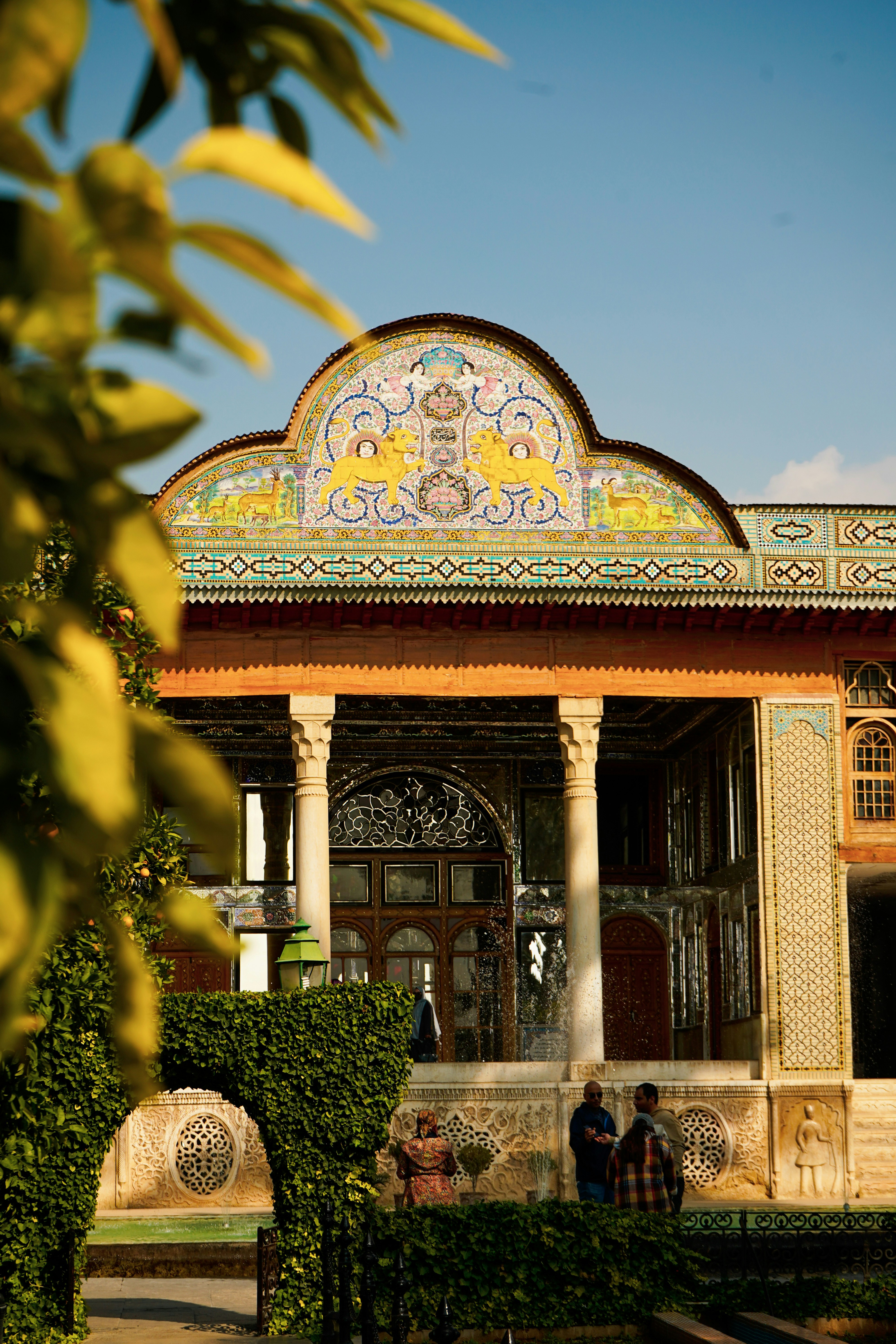 Intricate mirrored facade of a historic Persian building surrounded by lush greenery and visitors enjoying the serene atmosphere.