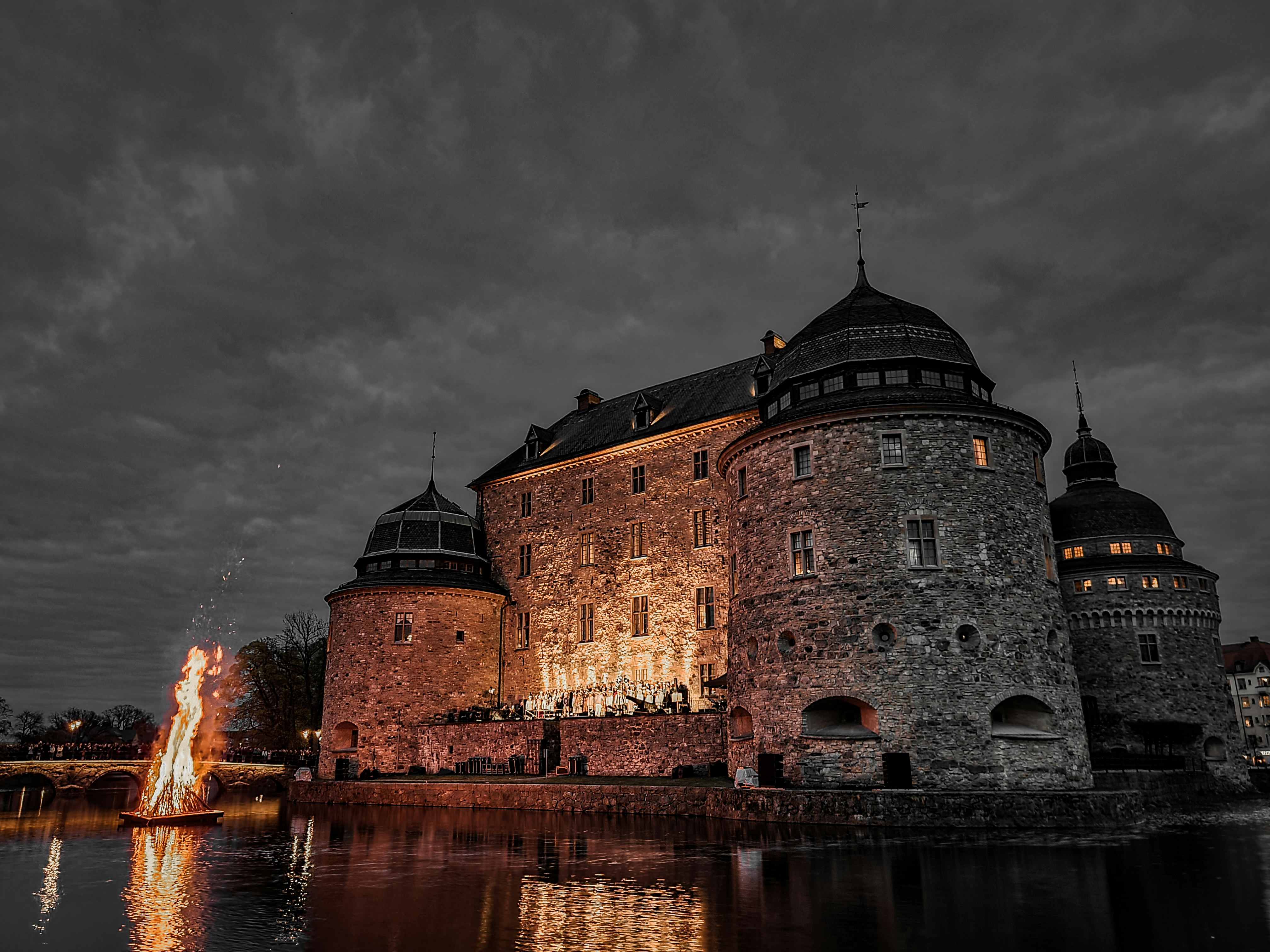 A majestic stone castle illuminated against a moody sky, with a fiery display reflecting in the water nearby.