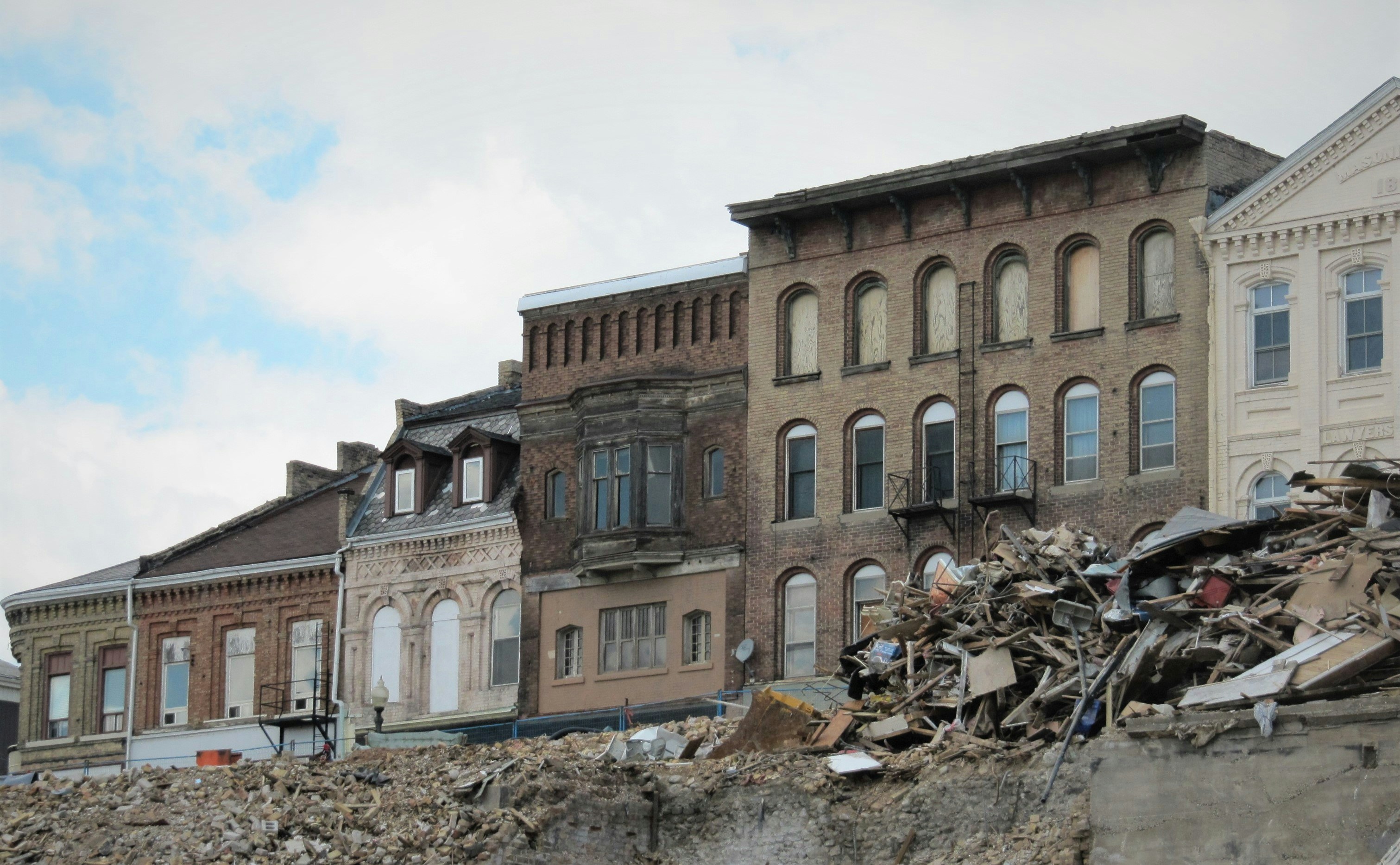 Historic buildings stand beside a pile of rubble under a cloudy sky.