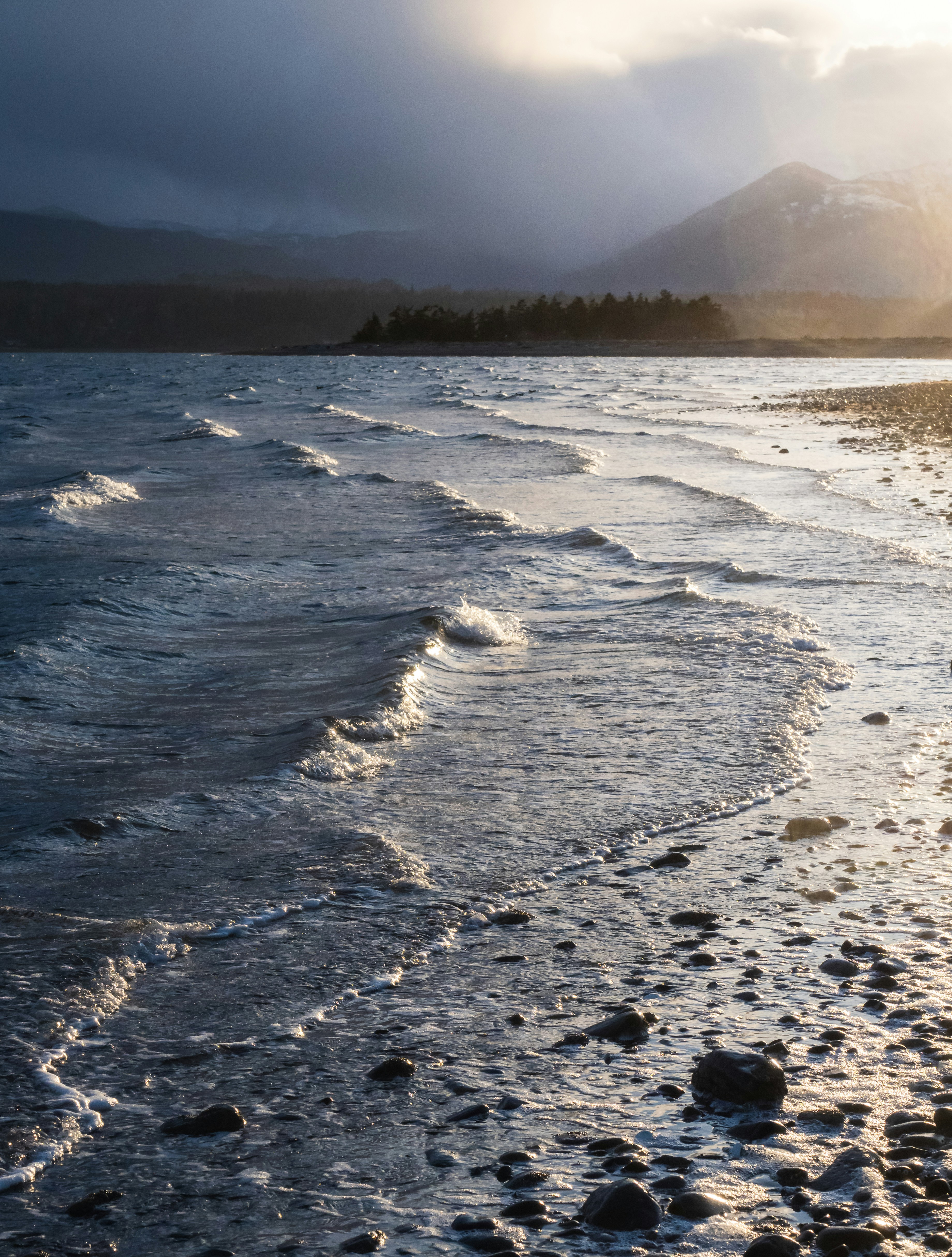 Gentle waves lap against a rocky beach under a moody sky, with distant mountains shrouded in mist. The scene captures the tranquil essence of nature's ebb and flow.