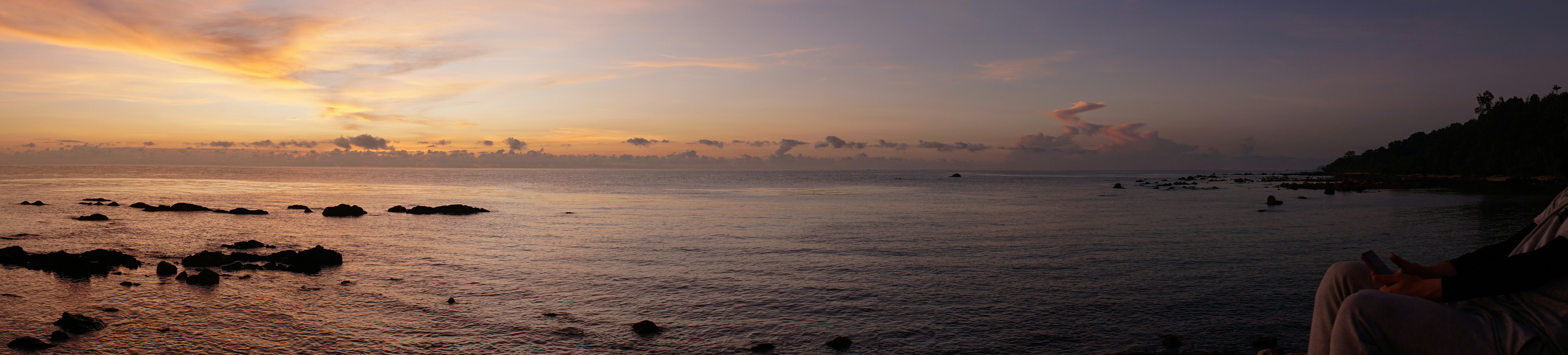 Calm ocean at sunrise with soft pastel skies and scattered rocks along the shoreline.