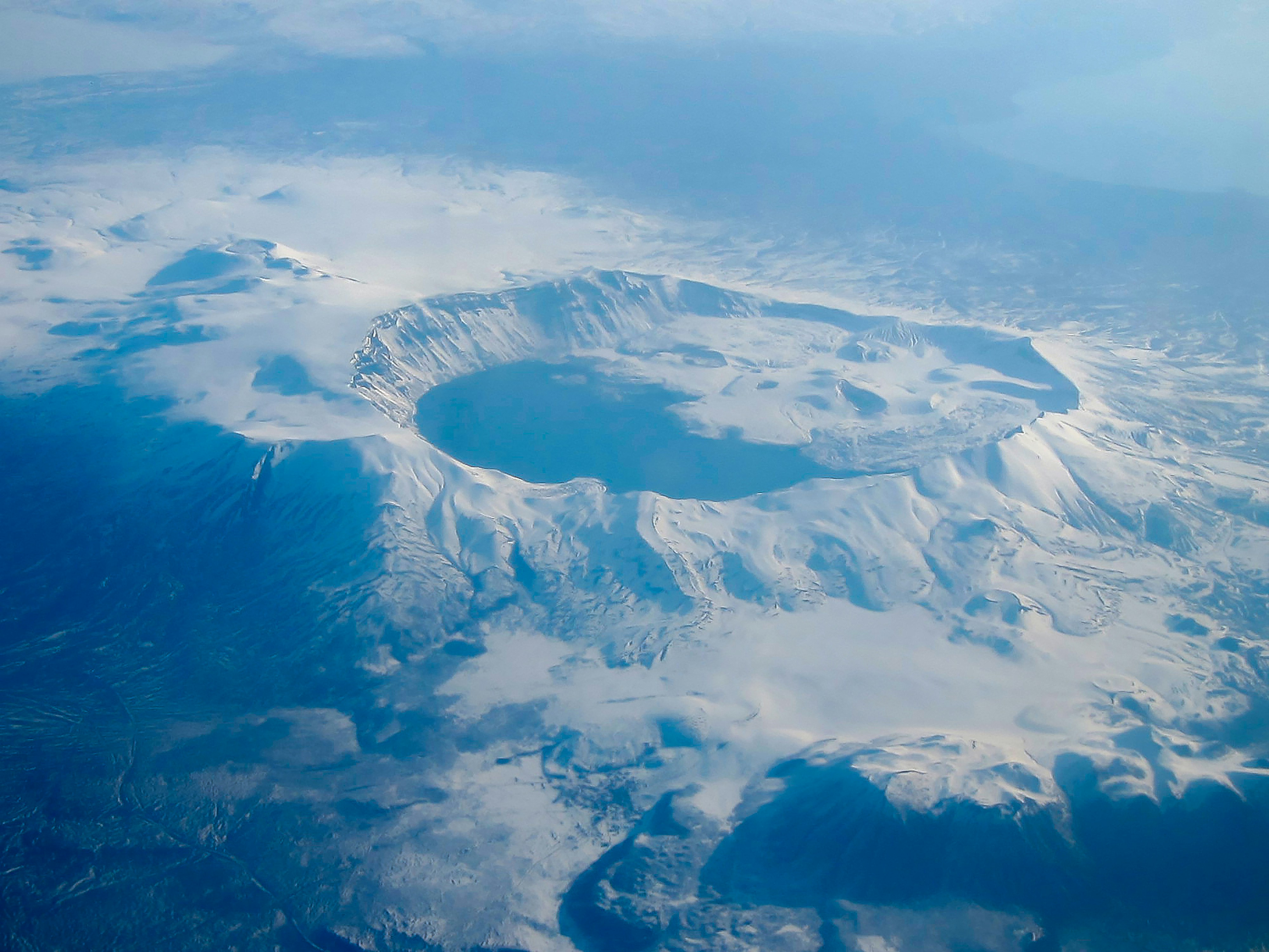 Aerial photograph of a colossal ice crater carved into a glacier, ringed by snow and ridges. The scene emphasizes the contrast between the dark crater interior and pale outer terrain.