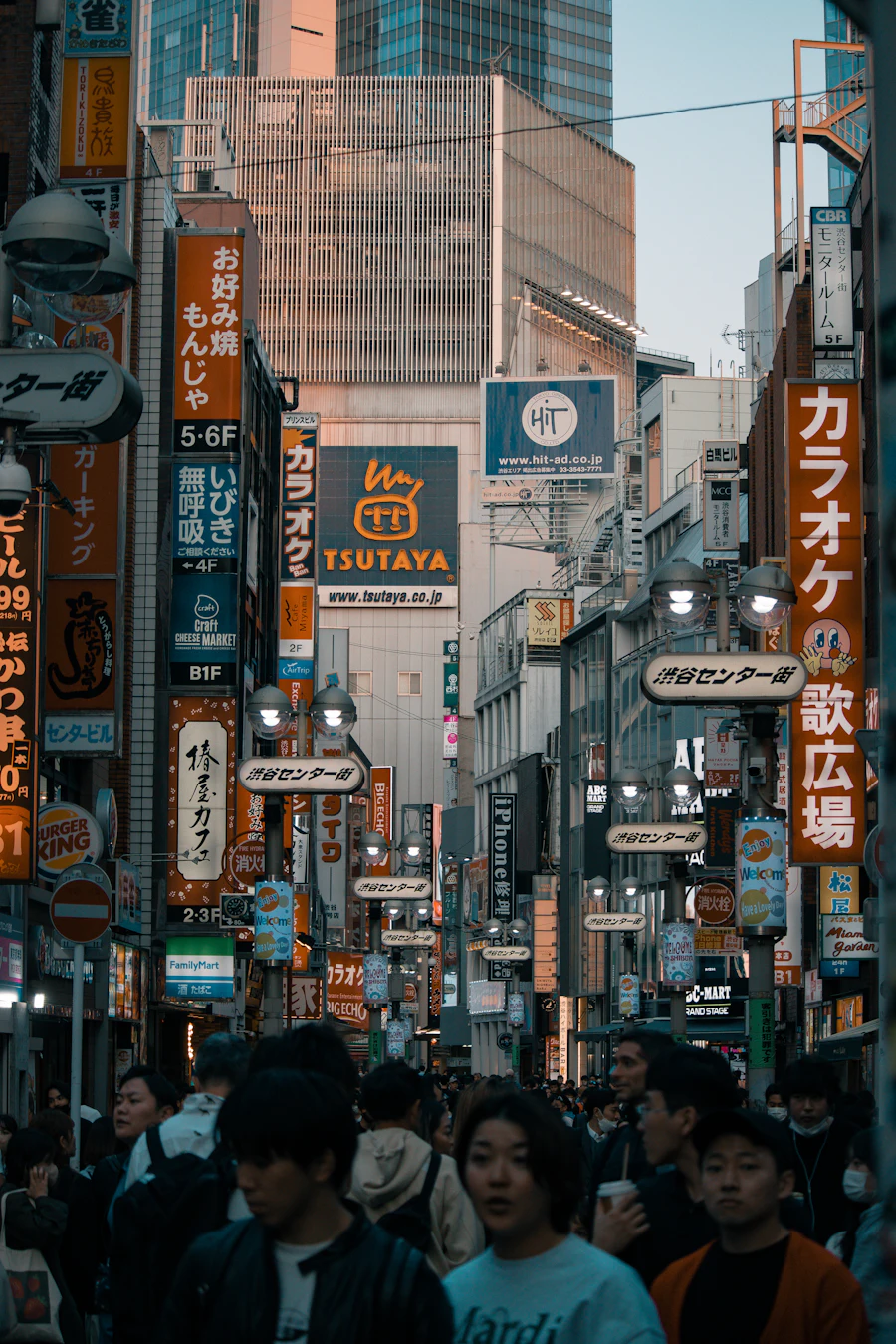 Crowded Tokyo shopping street packed with vertical signs, glowing storefronts, and pedestrians under tall buildings. Tsutaya and karaoke signs dominate the dense urban scene.