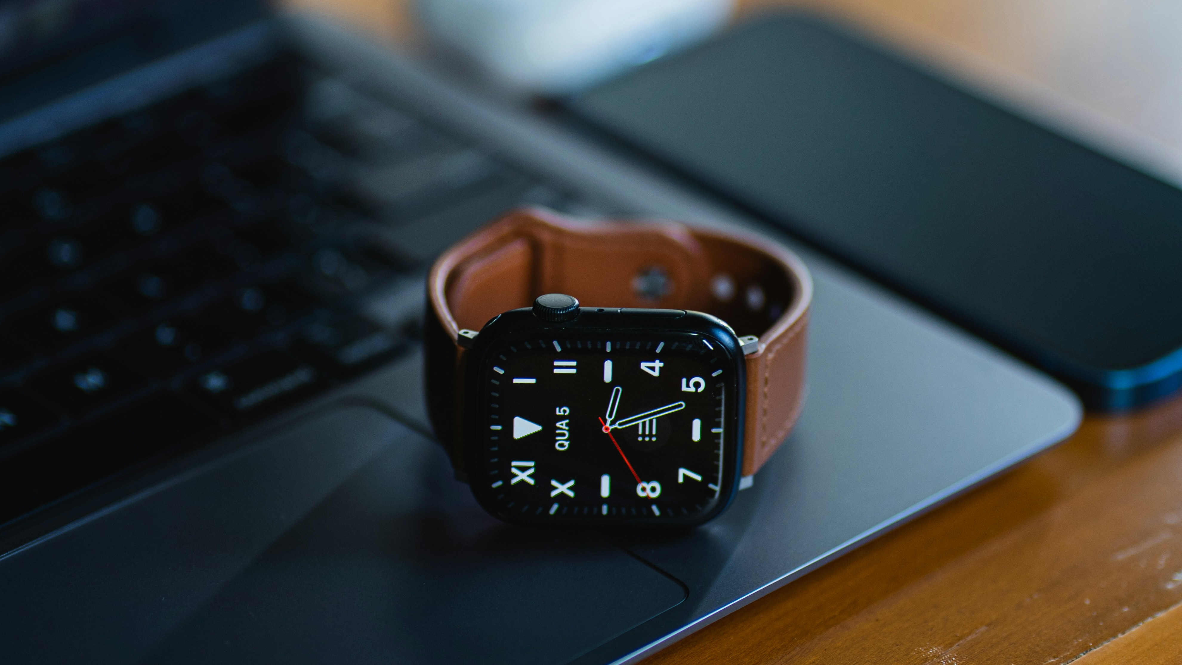 Smartwatch with a brown leather band resting on a laptop keyboard, beside a smartphone on a wooden desk.