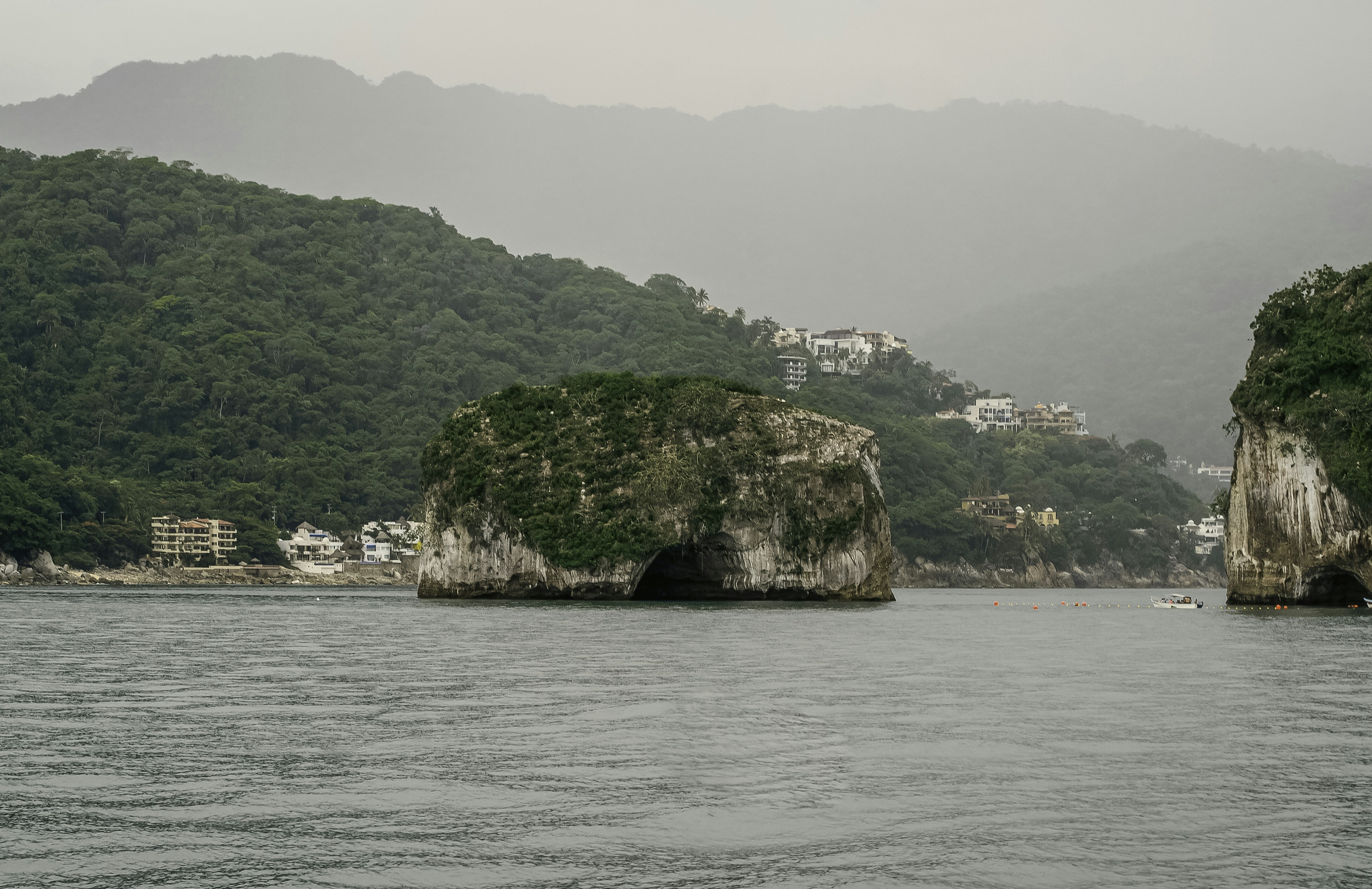 Lush green hills rise above the tranquil waters, framing a prominent rocky outcrop near the shoreline. A coastal village nestles in the background, hinting at human presence amidst nature's grandeur.