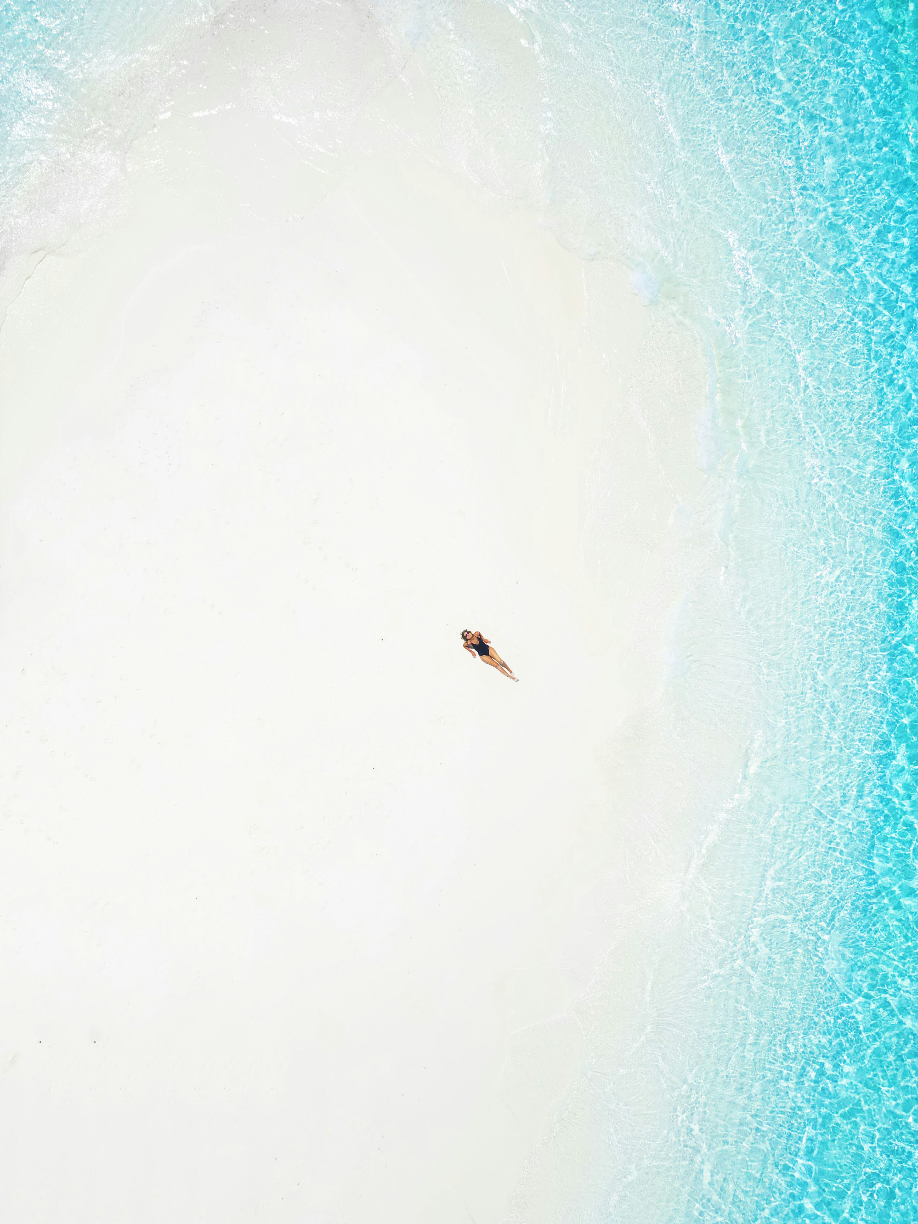 Aerial photograph of a lone figure on a stark white sandbar bordered by turquoise water. The composition highlights isolation against the vivid contrast of beach and sea.
