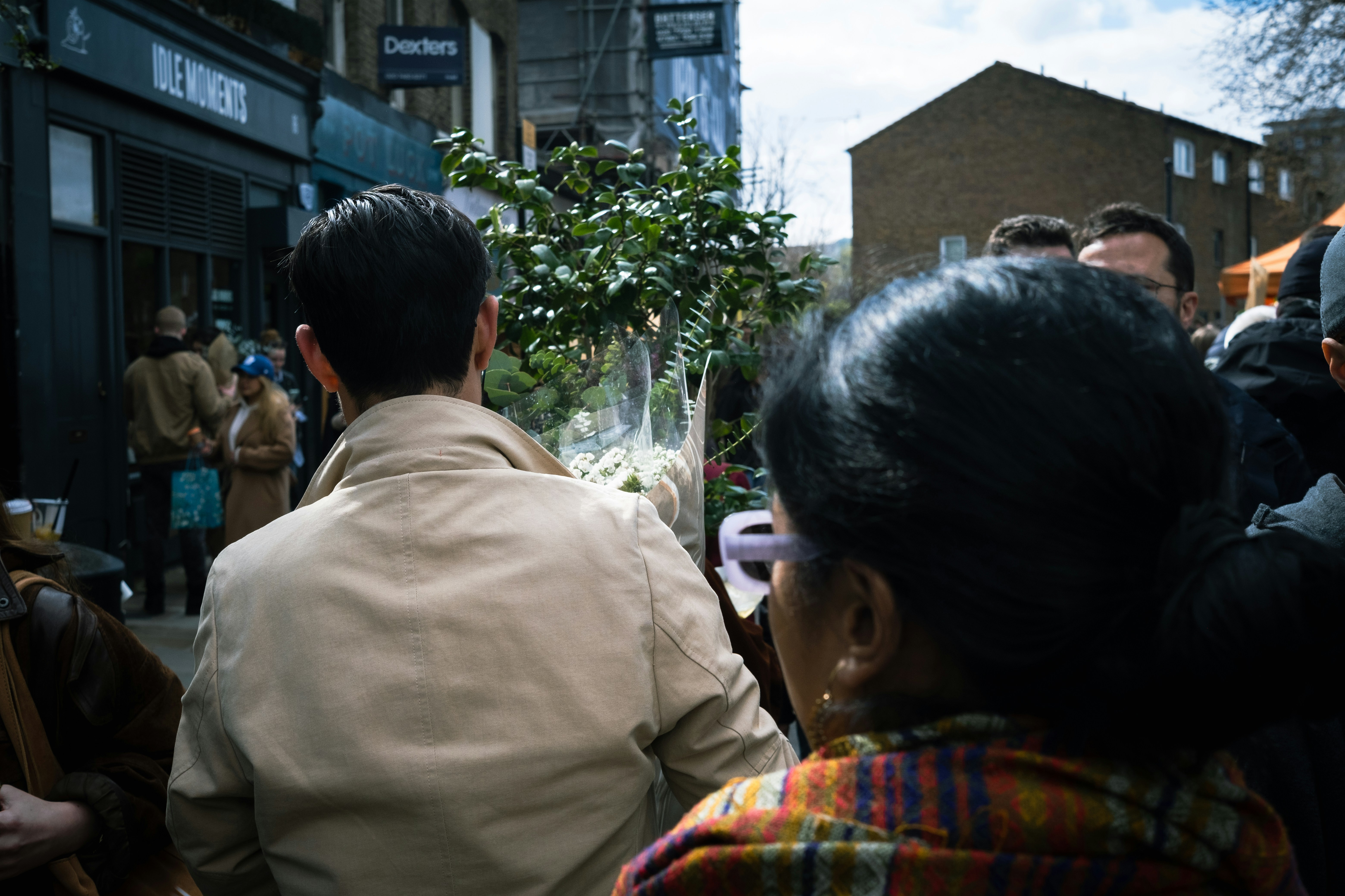 A person in a beige coat stands amidst a bustling street scene, with greenery and onlookers creating a lively atmosphere.