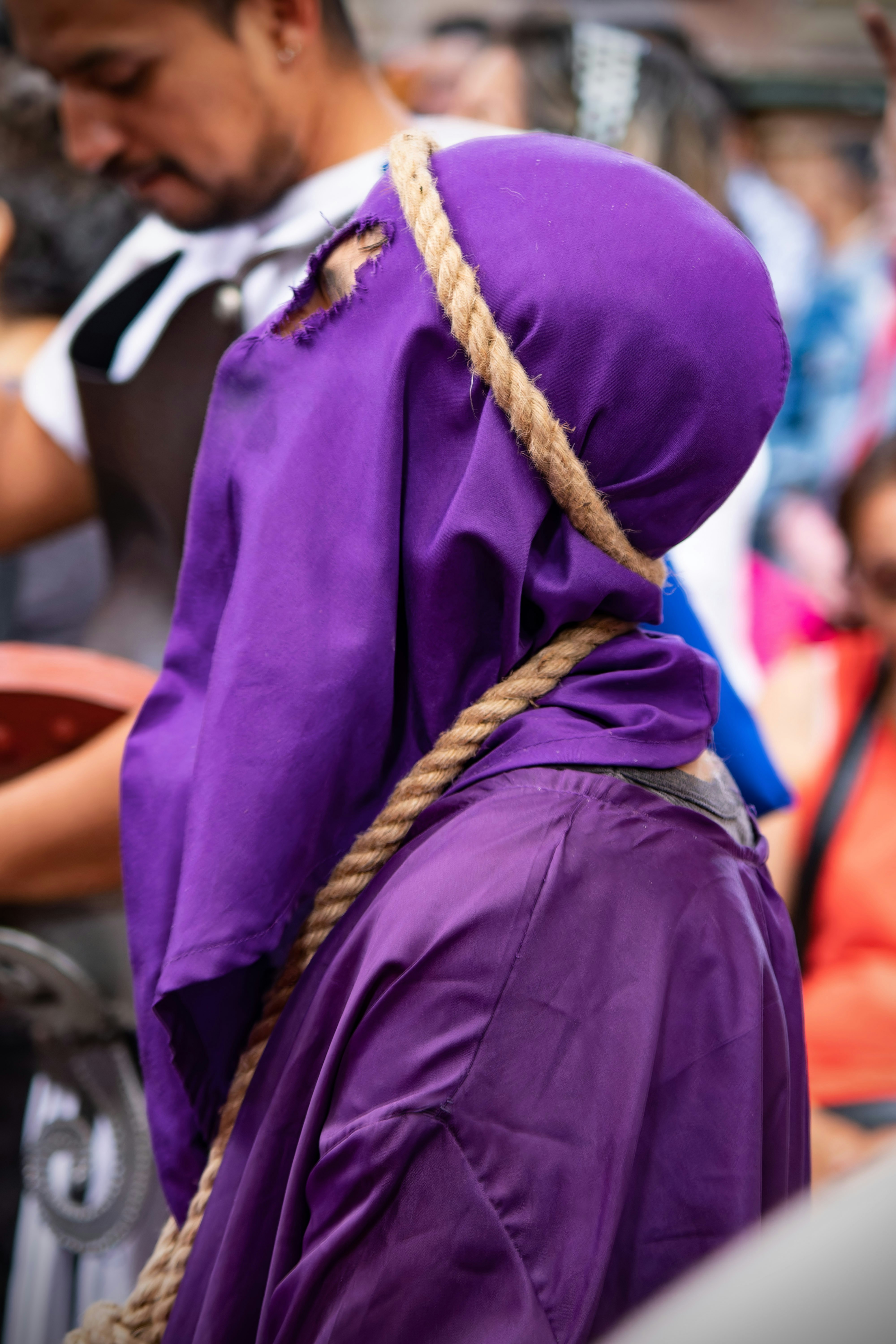 Person in purple hooded robe with rope detail, amidst a colorful crowd.